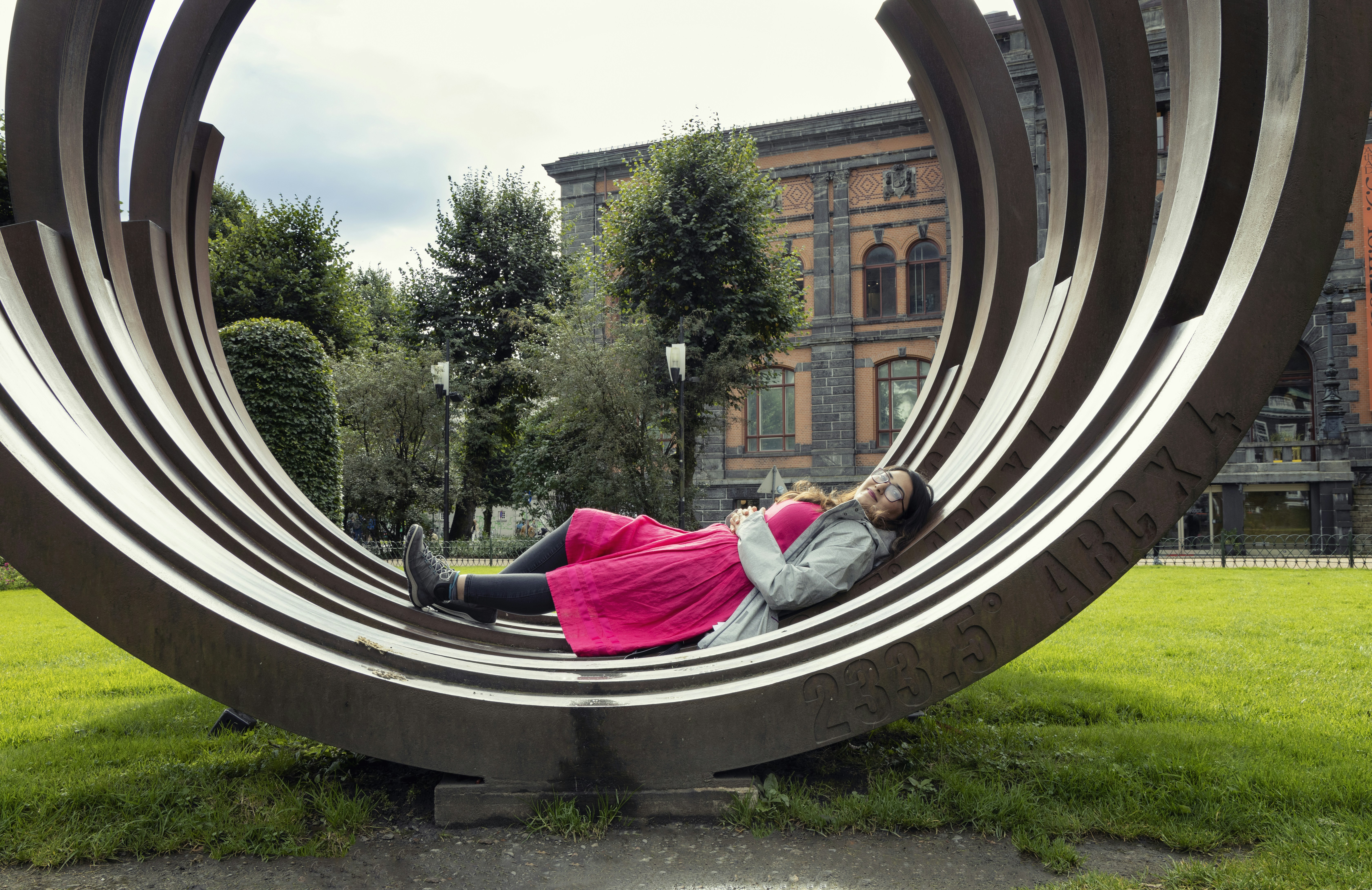 A photograph of a person in a bright pink dress reclining inside concentric steel rings in a park, with a brick building and green lawn in the background.