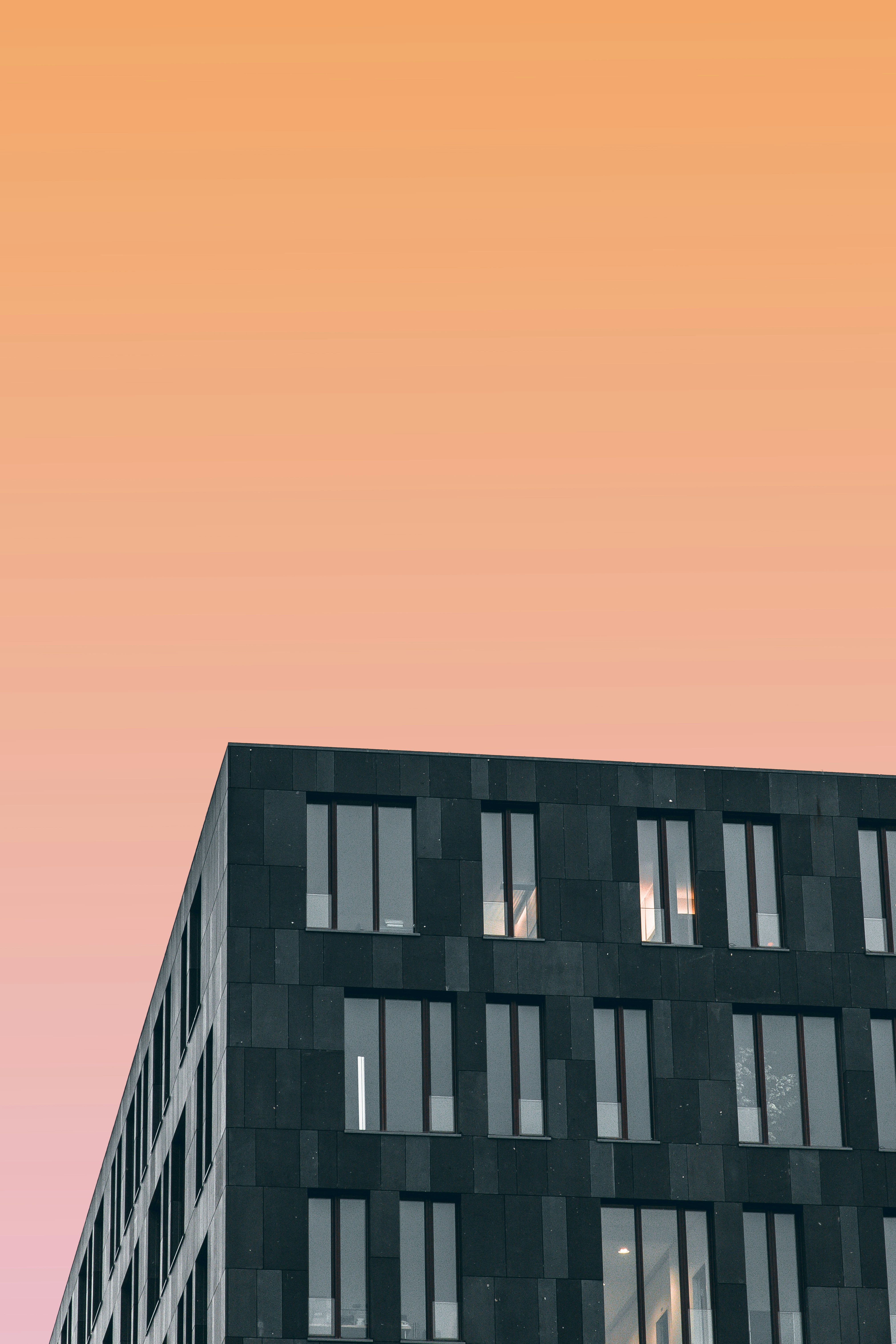 Contemporary building with a textured black facade and large windows, set against a gradient orange sky.