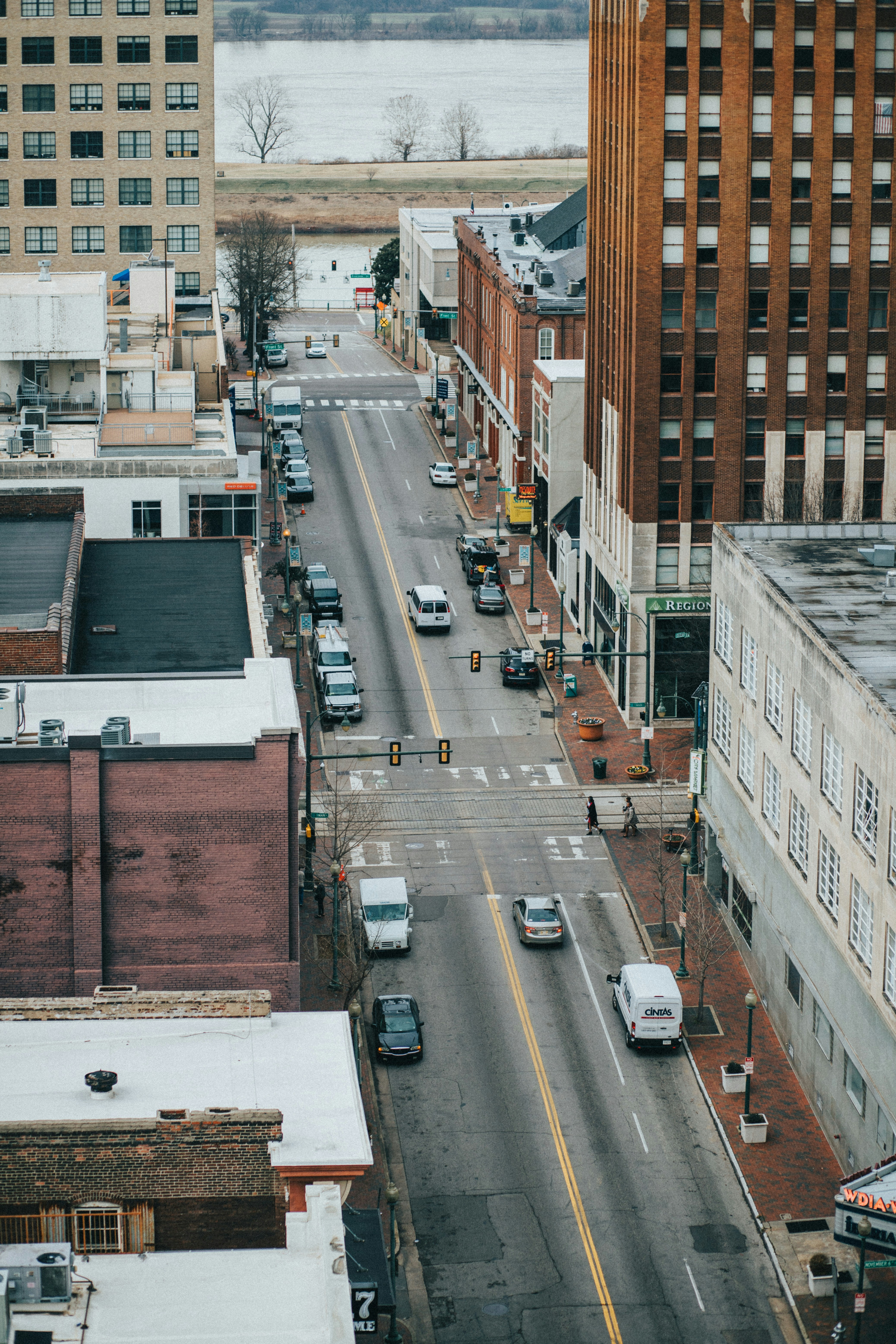 Aerial view of a bustling city intersection, showcasing vehicles navigating the streets and buildings lining the roads. The river is visible in the background.