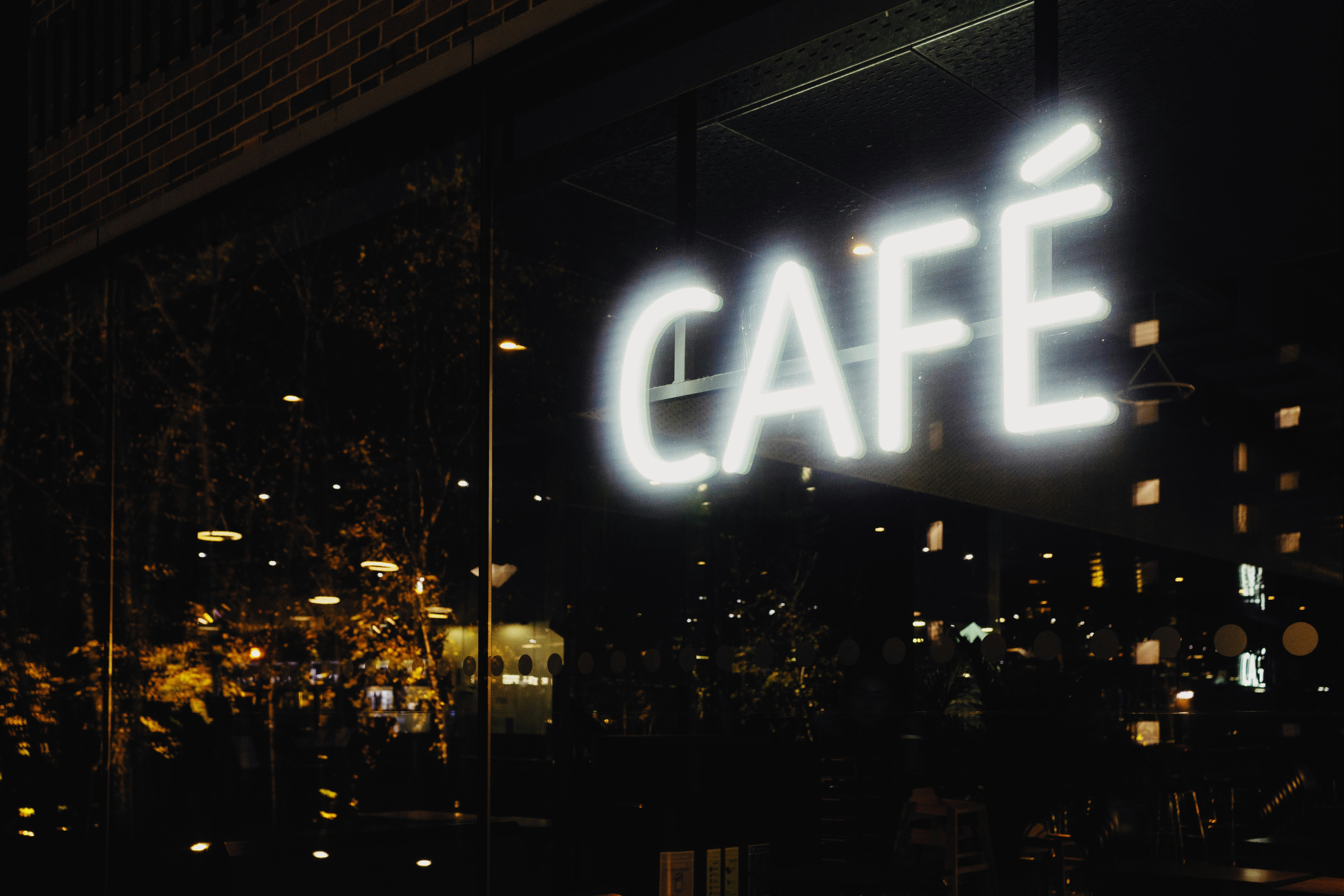 Bright neon sign reading 'CAFE' glows against a dark window, reflecting the warm ambiance of the café interior.