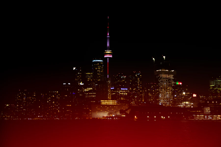 Night city skyline with the CN Tower glowing in the center and high-rise buildings lit around it. A dark foreground and red haze at the bottom frame the scene.