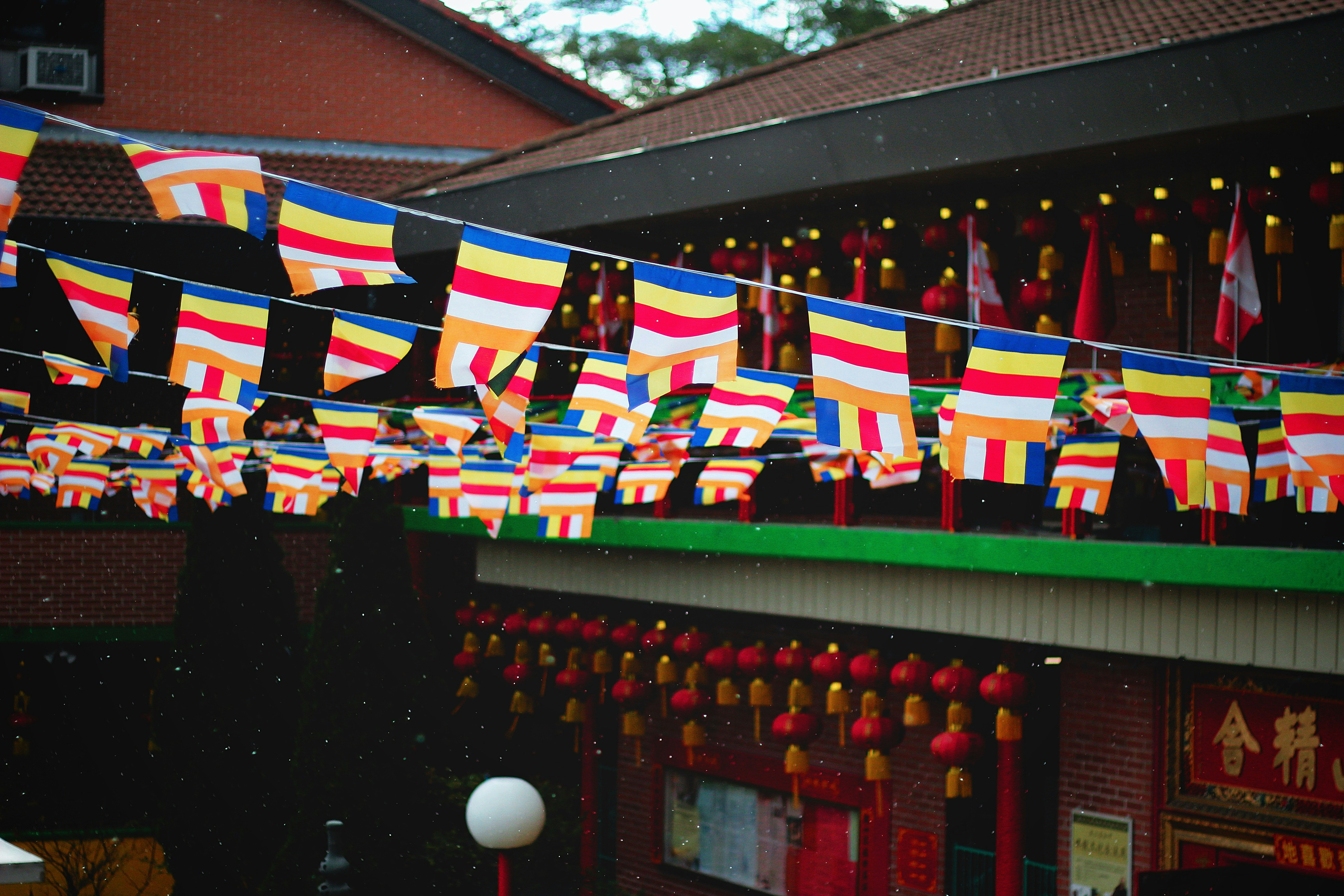 Colorful flags flutter above a Buddhist temple adorned with red lanterns during a gentle snowfall.