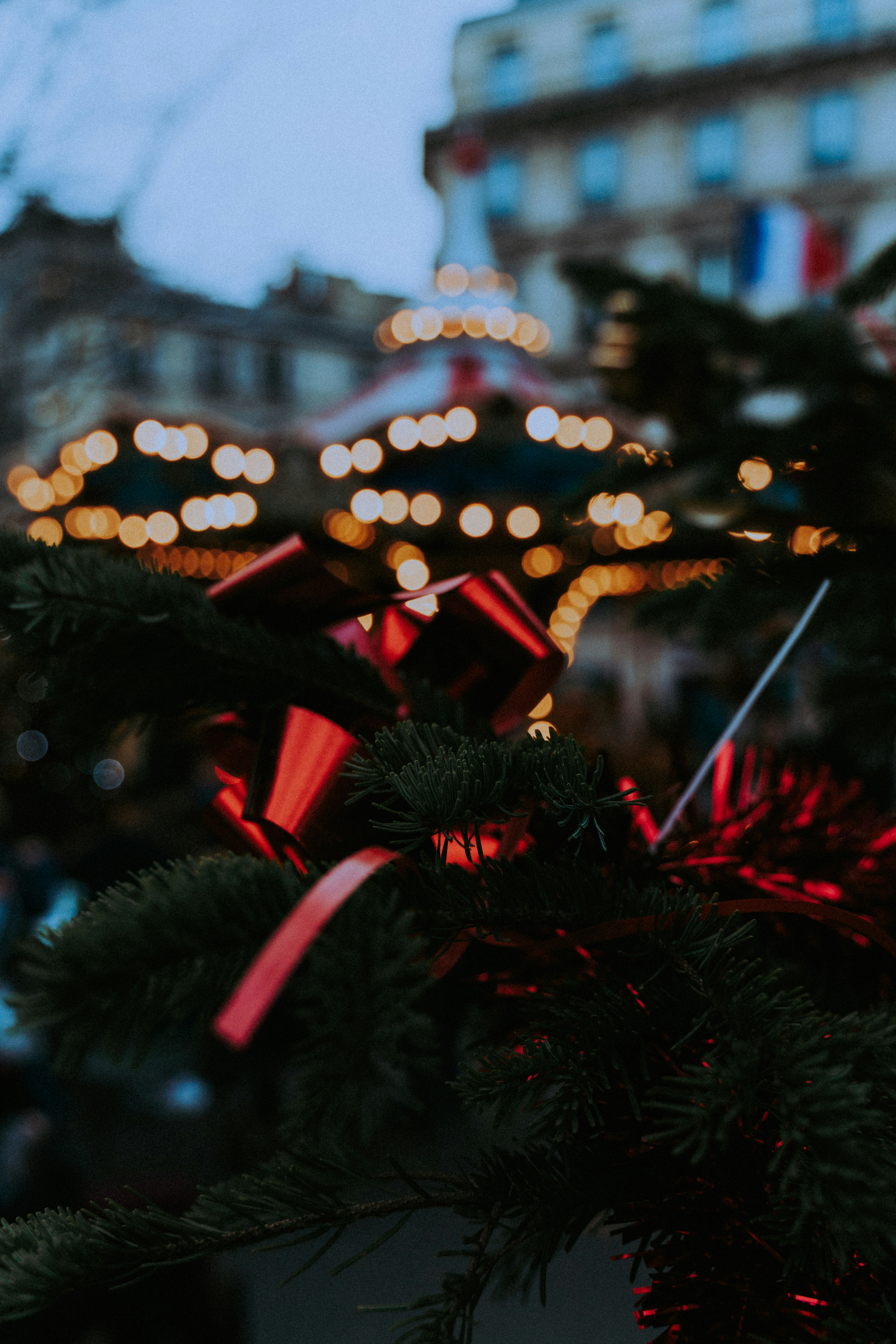 A close-up of festive decorations on an evergreen branch, with a blurred carousel illuminated by warm lights in the background.