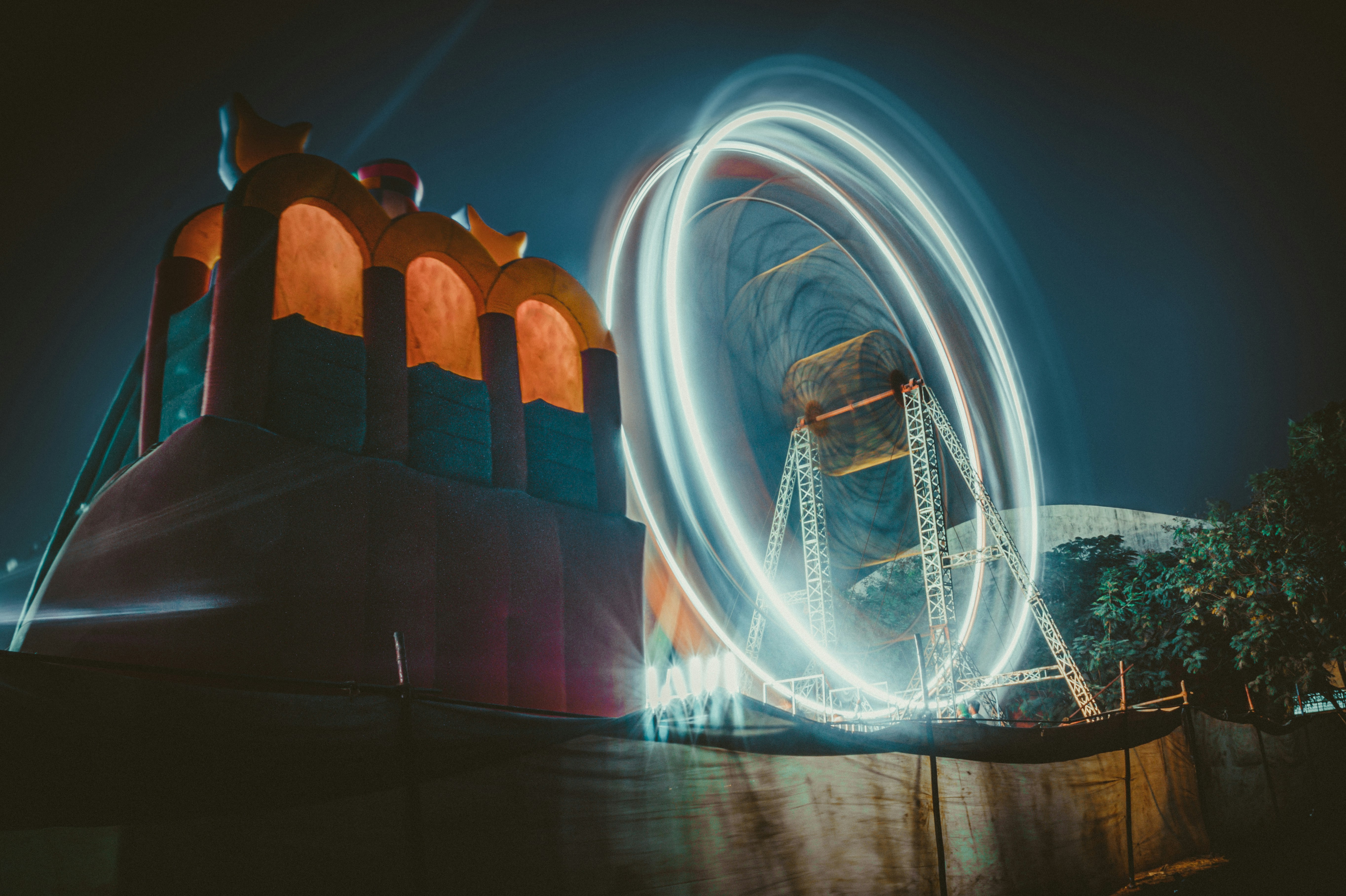 Long-exposure photograph of a spinning light ring at a night carnival beside a fortress-like, neon-lit structure.