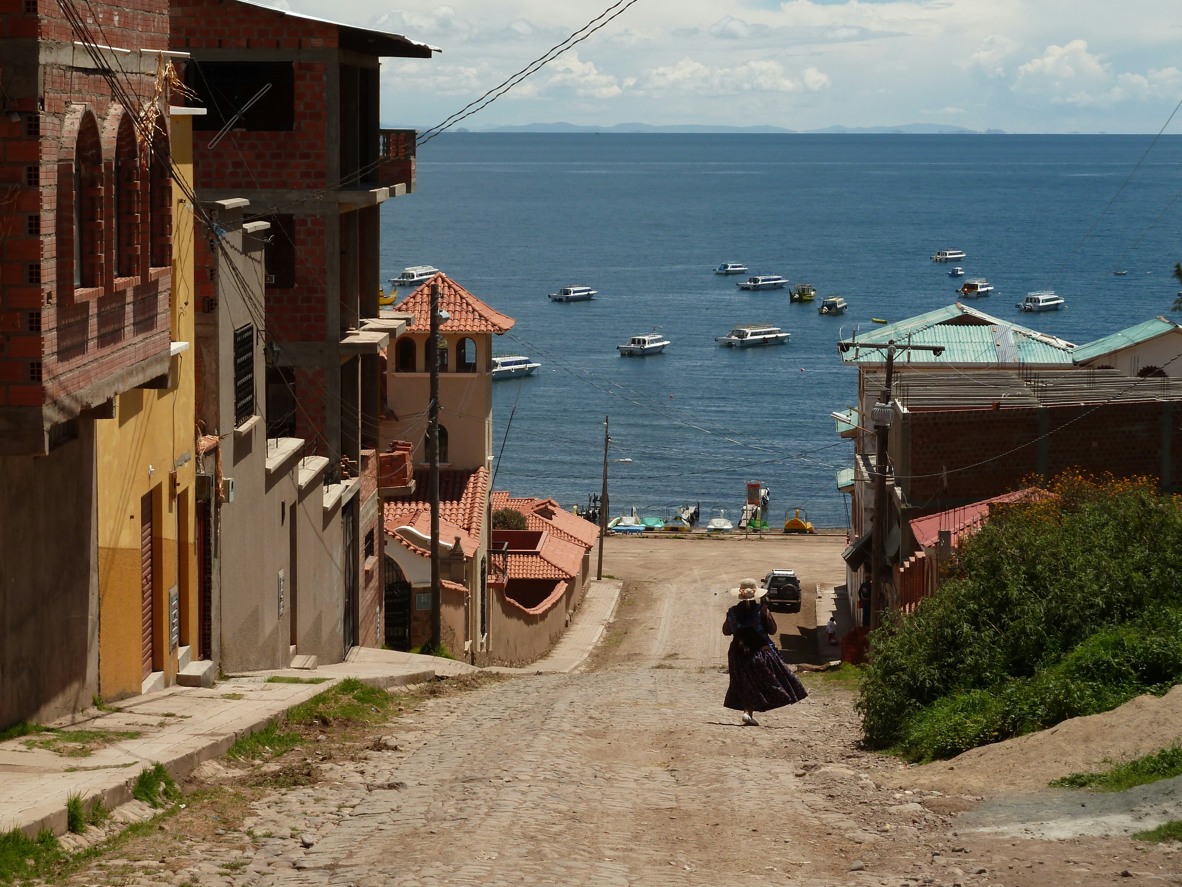 A woman in traditional attire walks down a cobblestone street towards a tranquil lake dotted with boats, framed by colorful buildings and lush greenery.