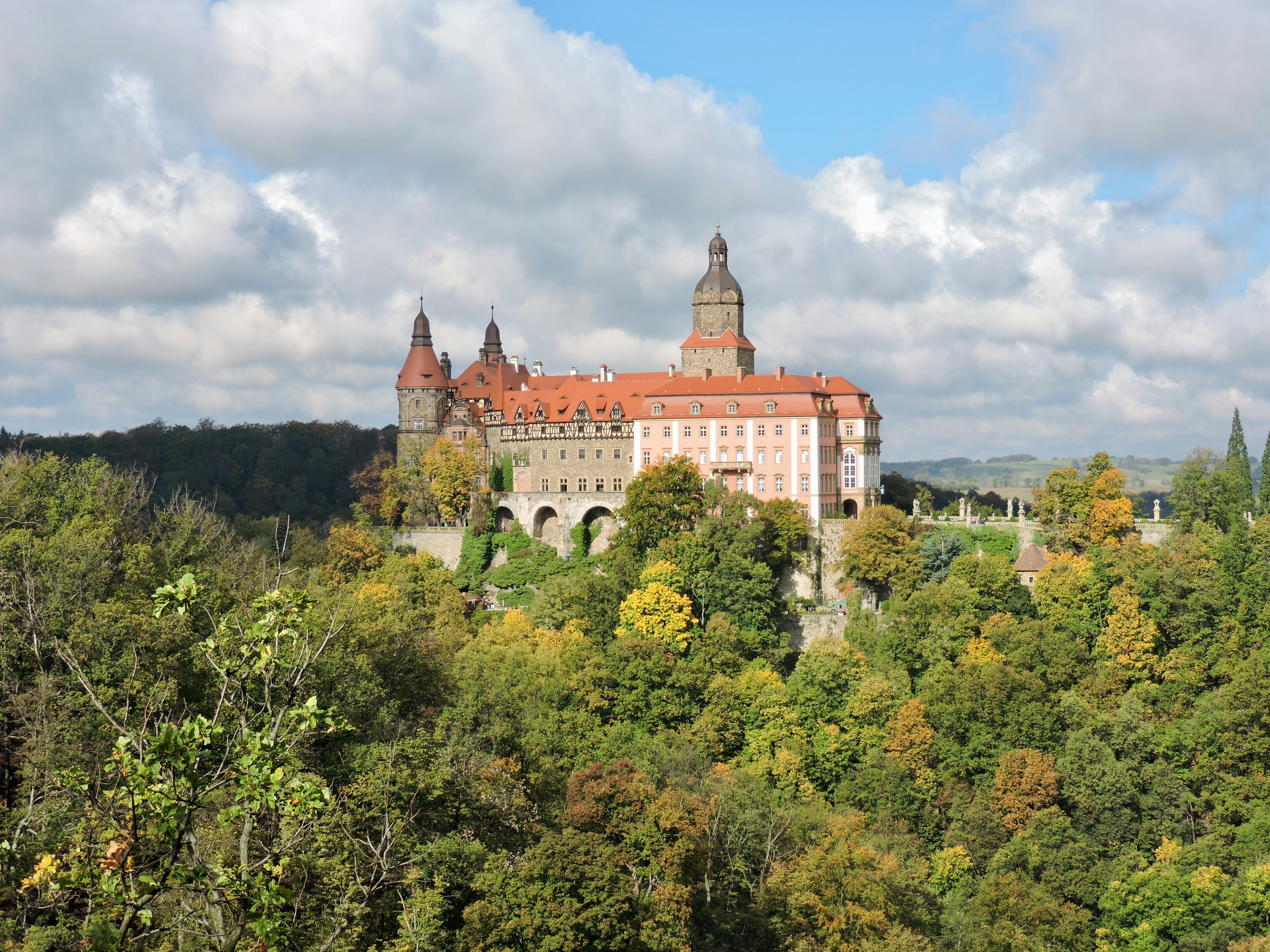 Historic castle perched on a lush hill surrounded by vibrant autumn trees under a partly cloudy sky.