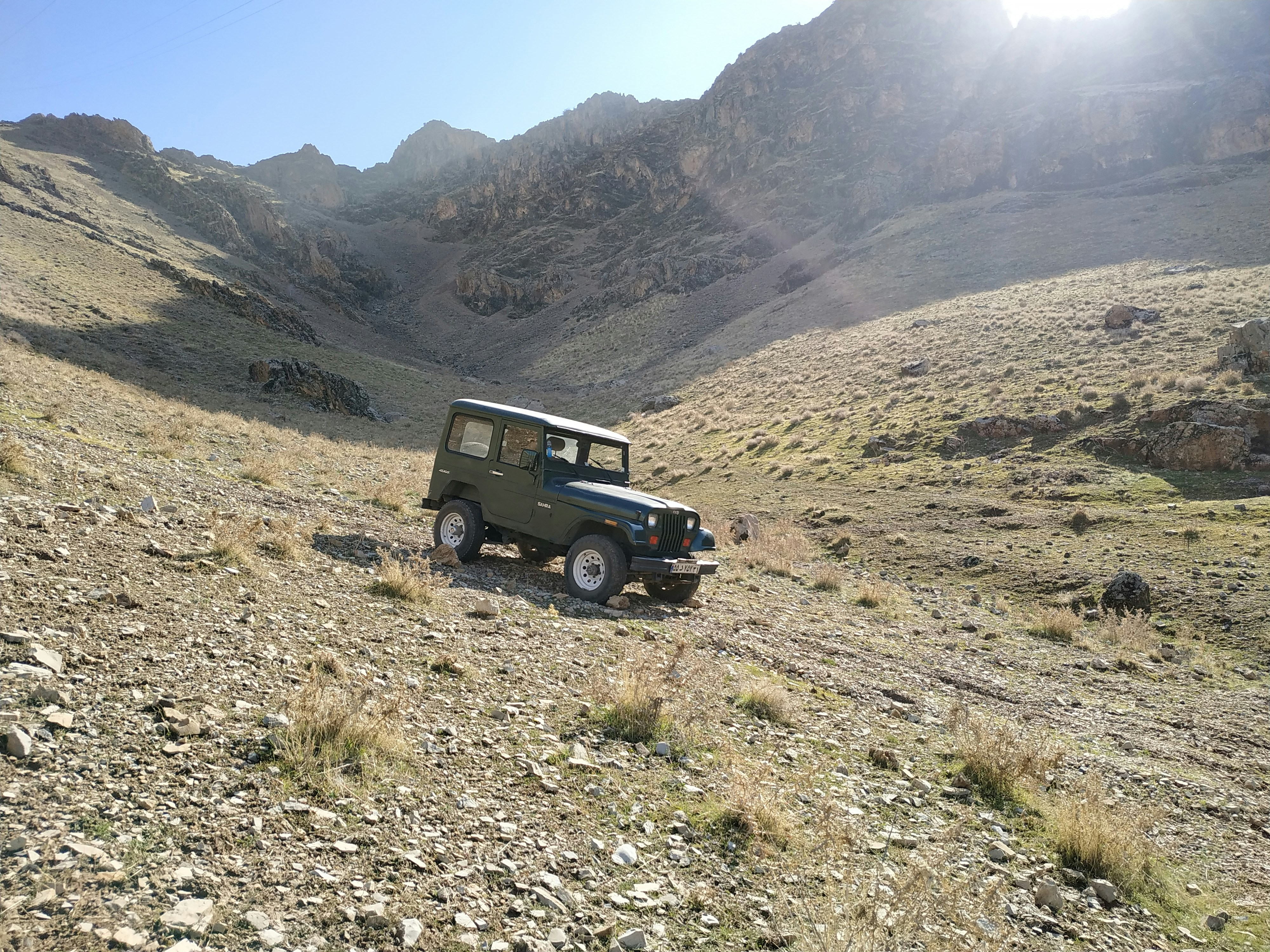 A green off-road vehicle navigating a rocky path in a mountainous landscape under a clear blue sky.