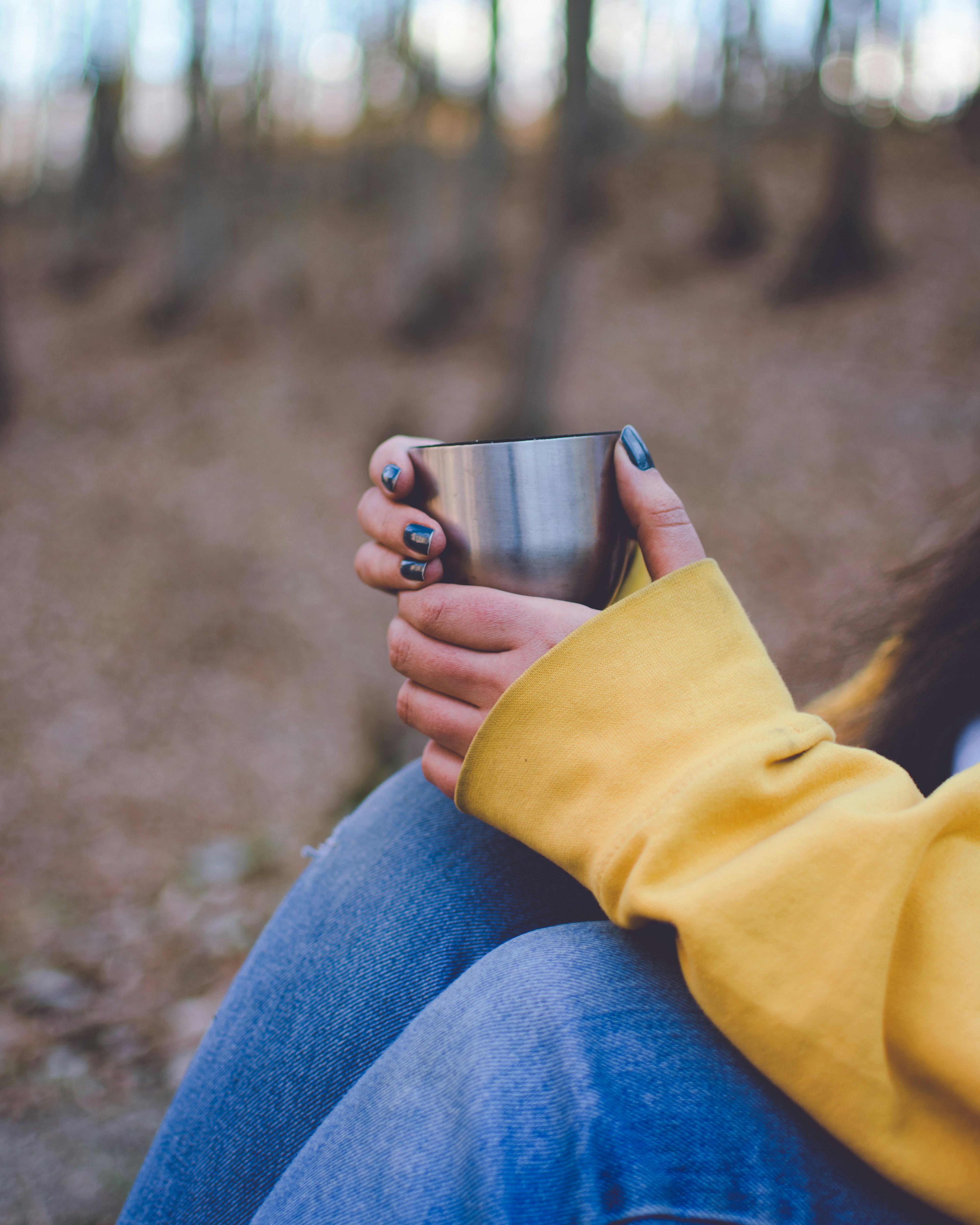 a girl holding a metal cup of tea in forest with blue jeans and yellow hoodie.