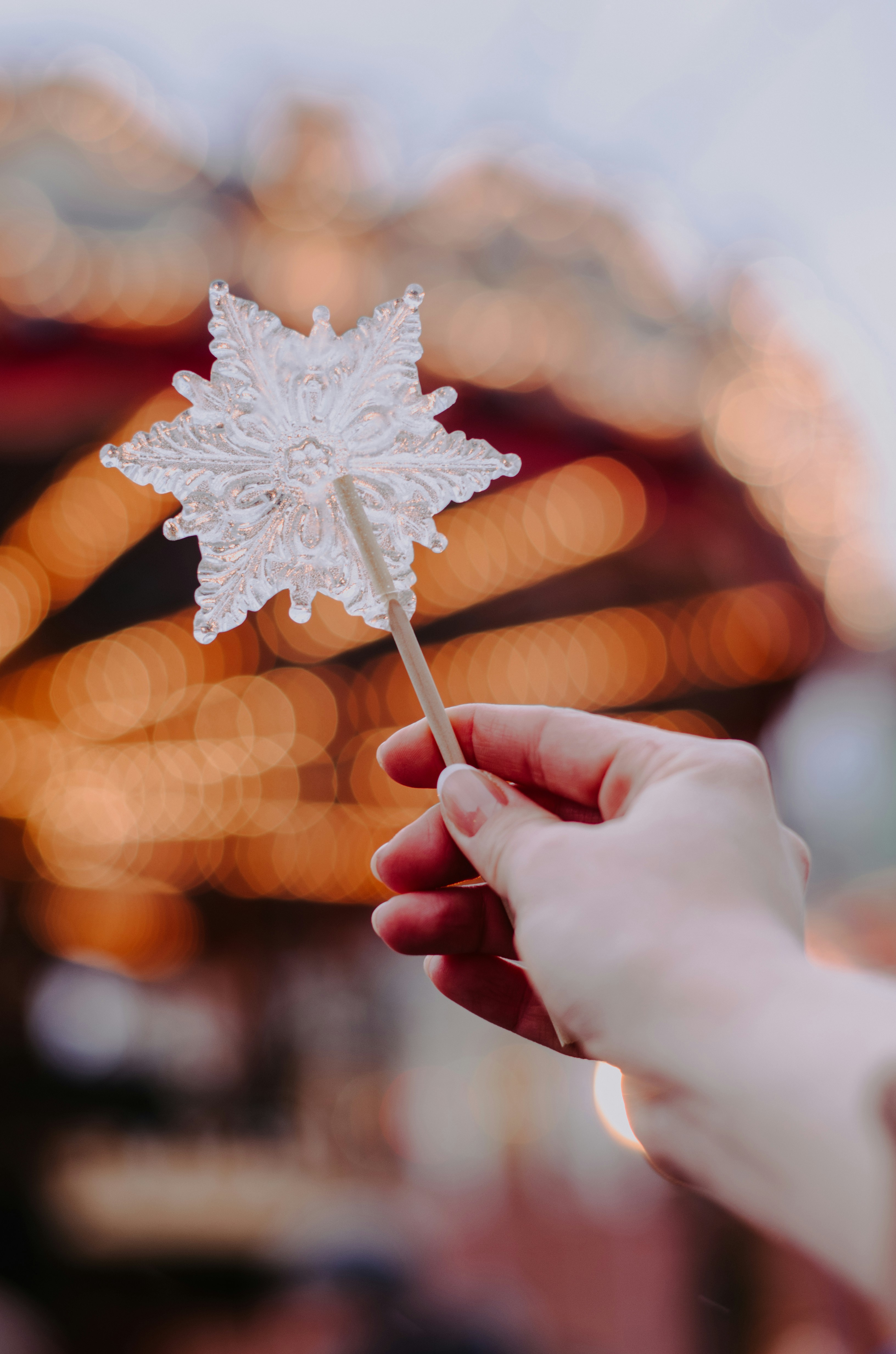 A frosted snowflake lollipop is held by a hand in sharp focus, with warm golden bokeh lights creating a soft backdrop.