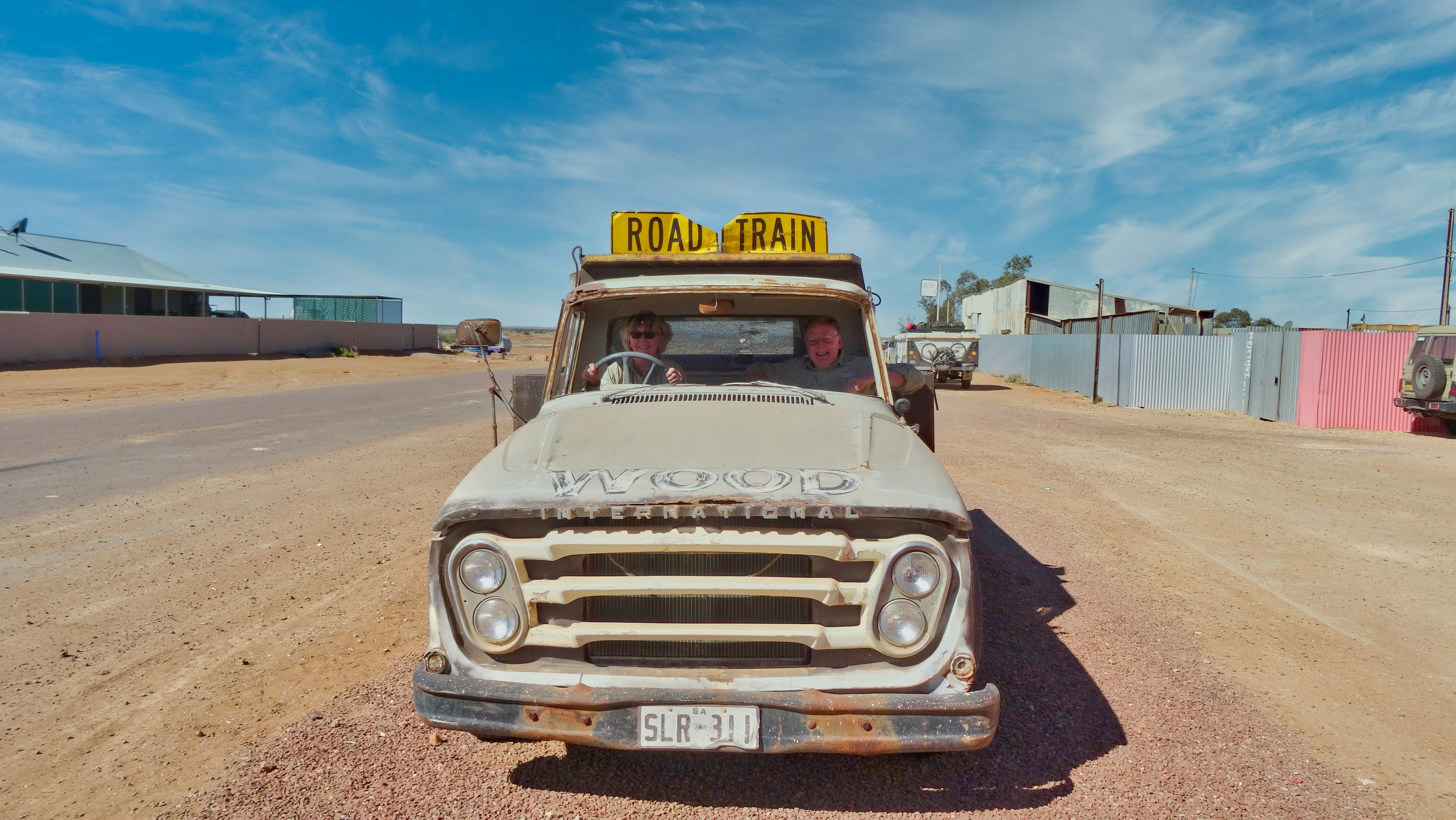 Rusty vintage truck centered on a dusty desert road beneath a bright blue sky, with a yellow ROAD TRAIN sign perched on its roof.