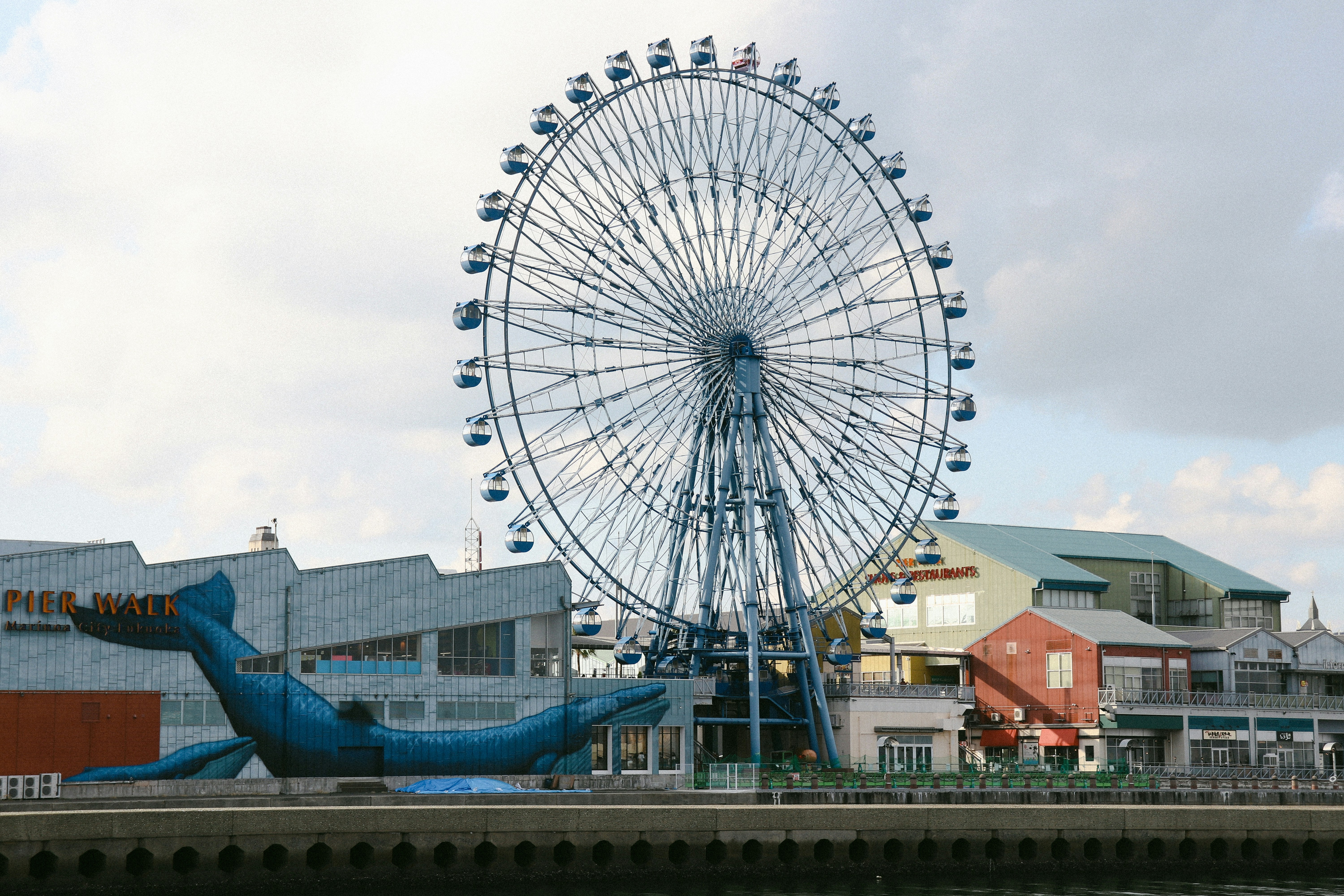 A giant Ferris wheel towers over a waterfront scene, accompanied by a striking whale mural on a building. The combination creates a playful atmosphere.