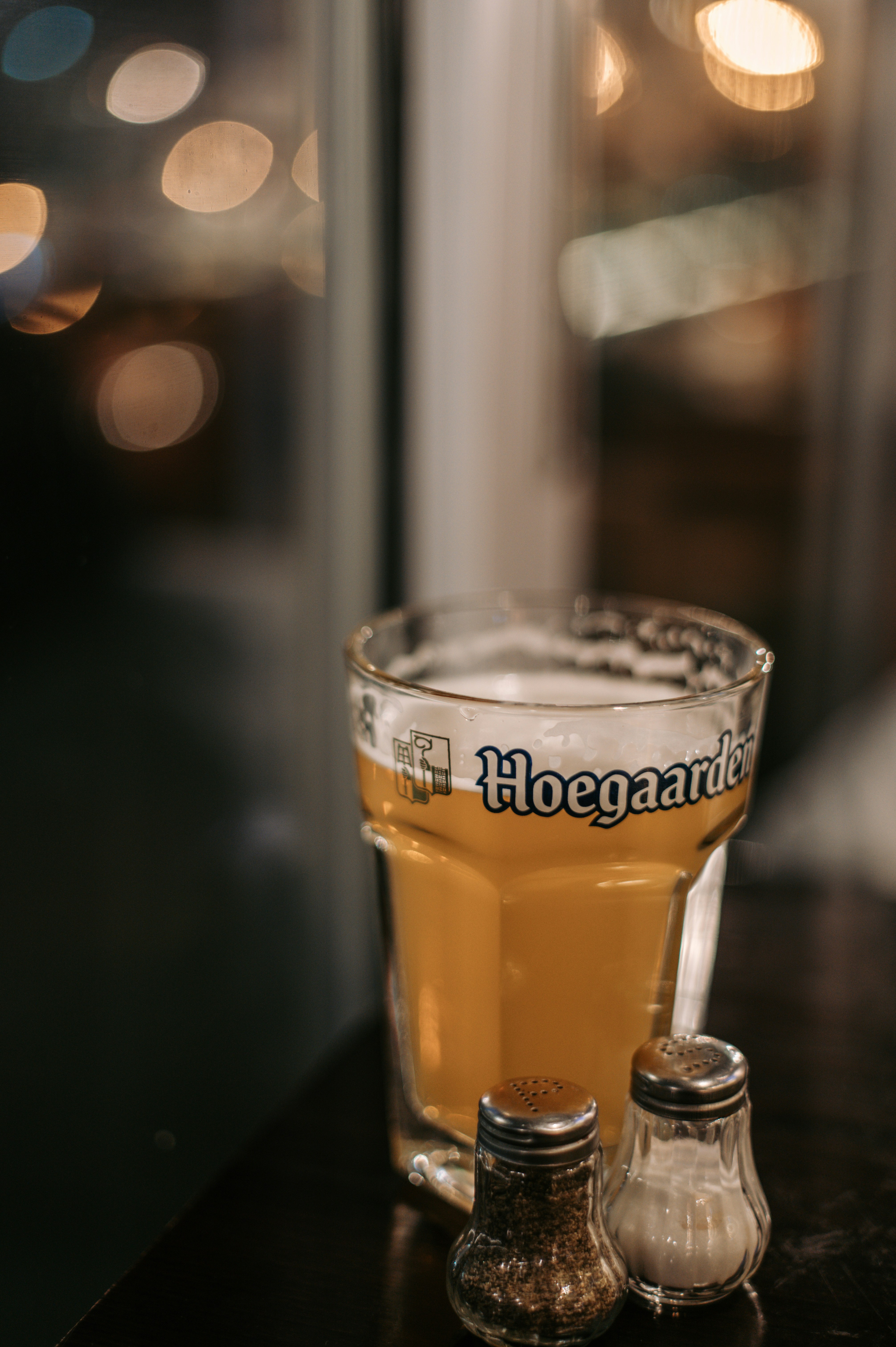 A glass of Hoegaarden beer sits on a table next to salt and pepper shakers, illuminated by soft background lights.