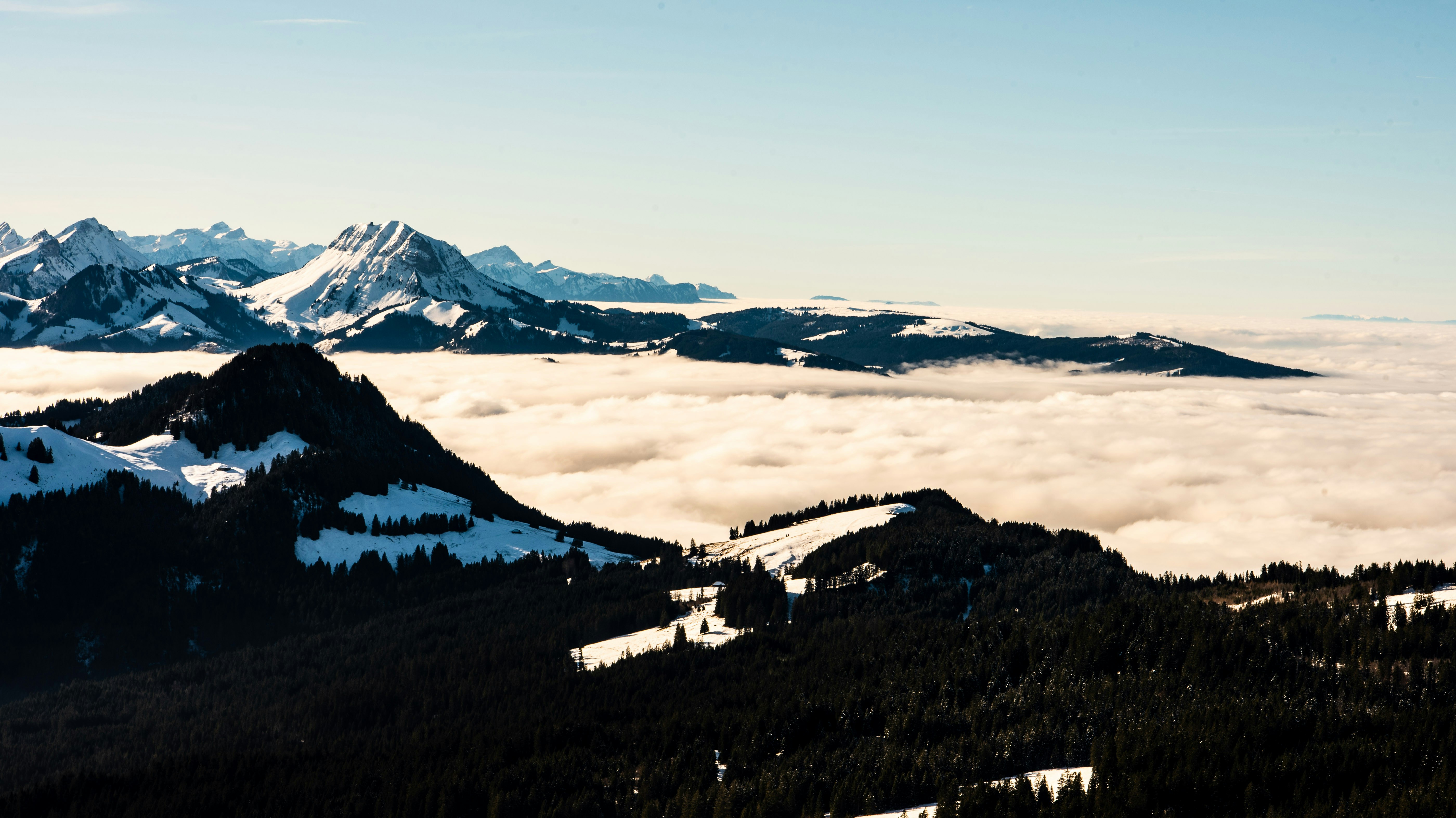 Snow-capped mountains rise above a sea of clouds, creating a serene landscape at dawn. The interplay of light and shadow highlights the rugged terrain.