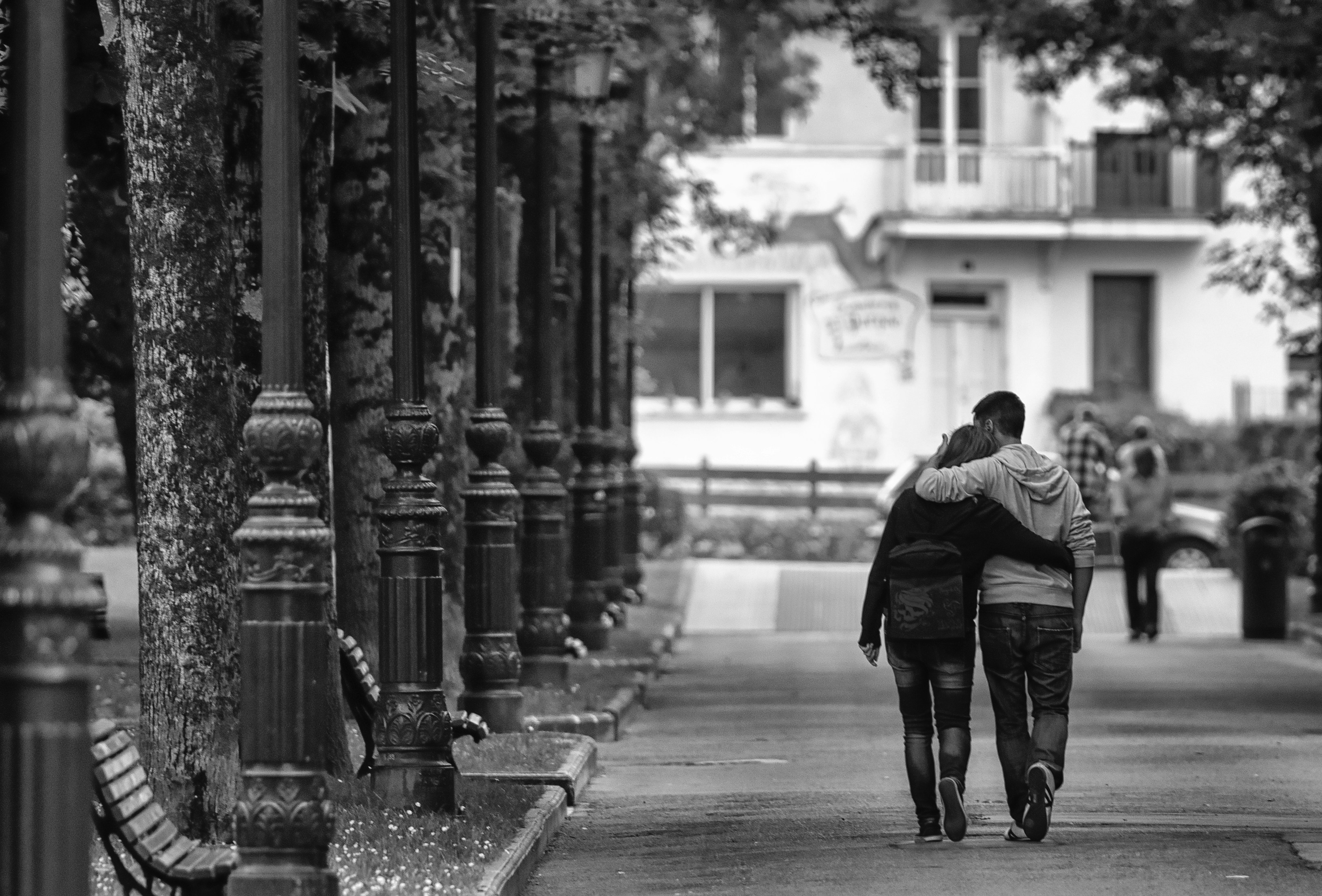 Couple walking closely together in a serene park, surrounded by historic lampposts and trees. A sense of intimacy permeates the scene.