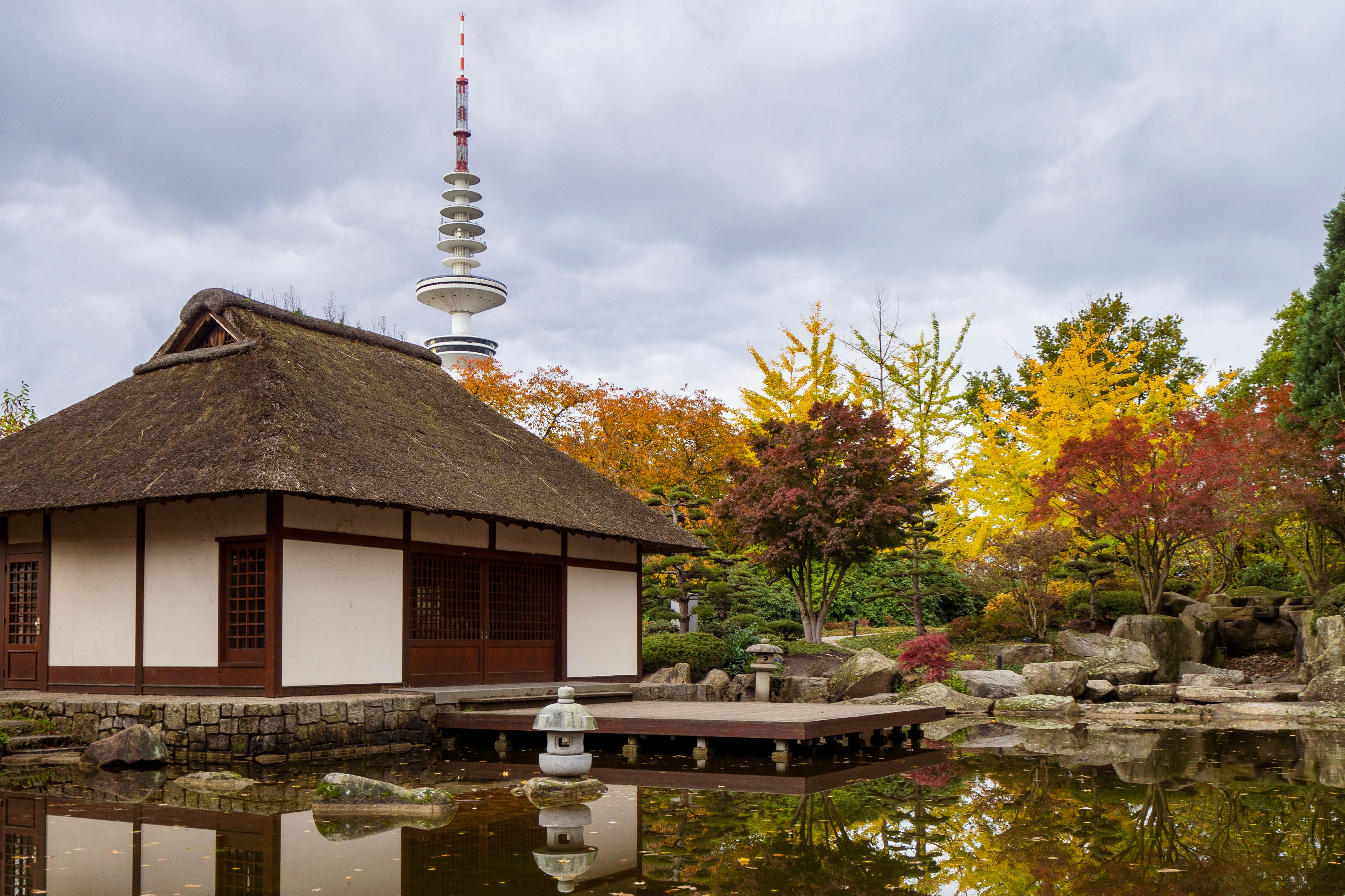 A traditional pavilion sits beside a calm pond, its reflection mirrored in the water as colorful autumn trees line the background; a serene garden scene captured near dusk.
