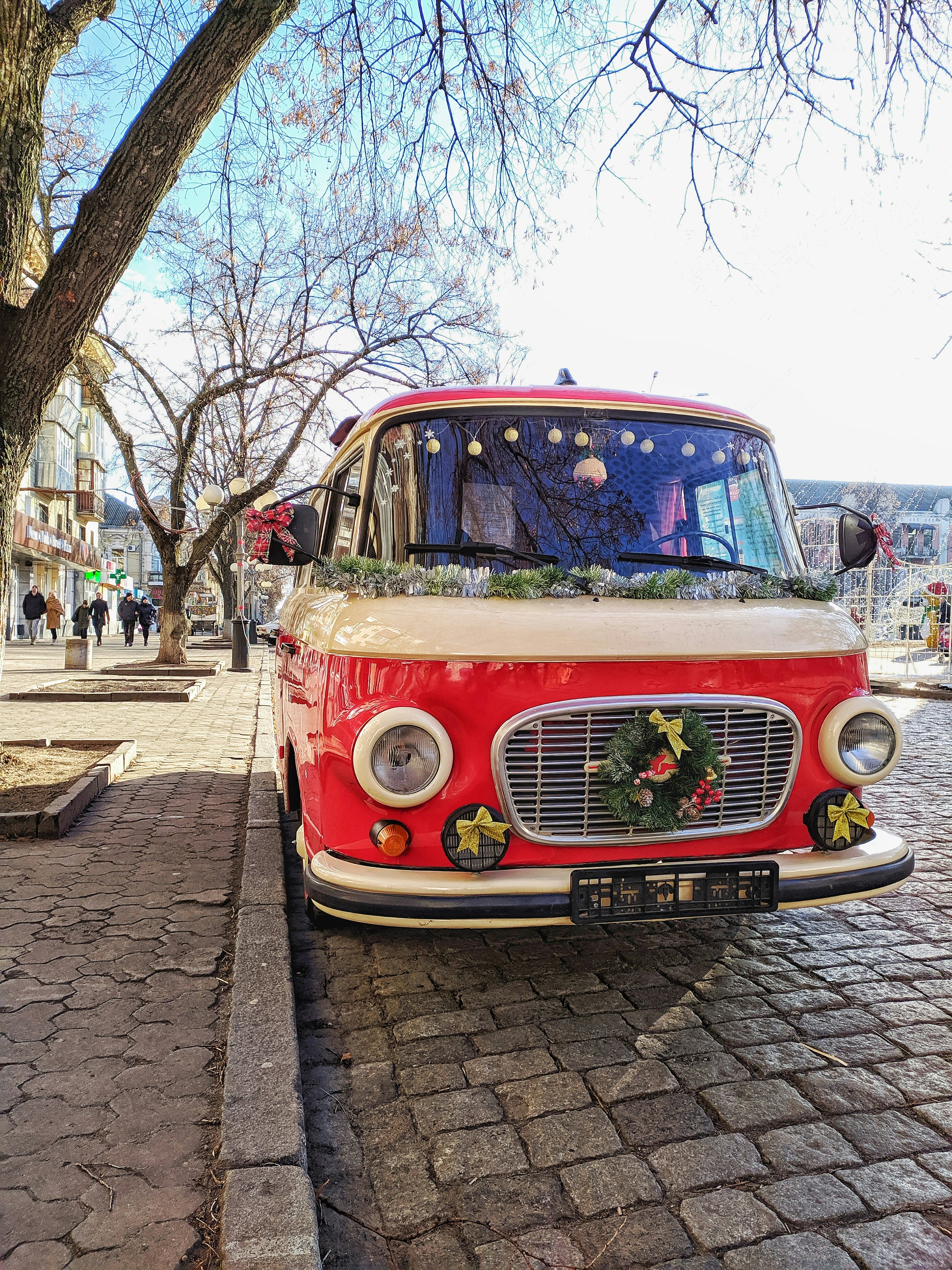 Front view of a red vintage van decorated with a wreath and garlands, parked on a cobblestone street with a bustling city backdrop.