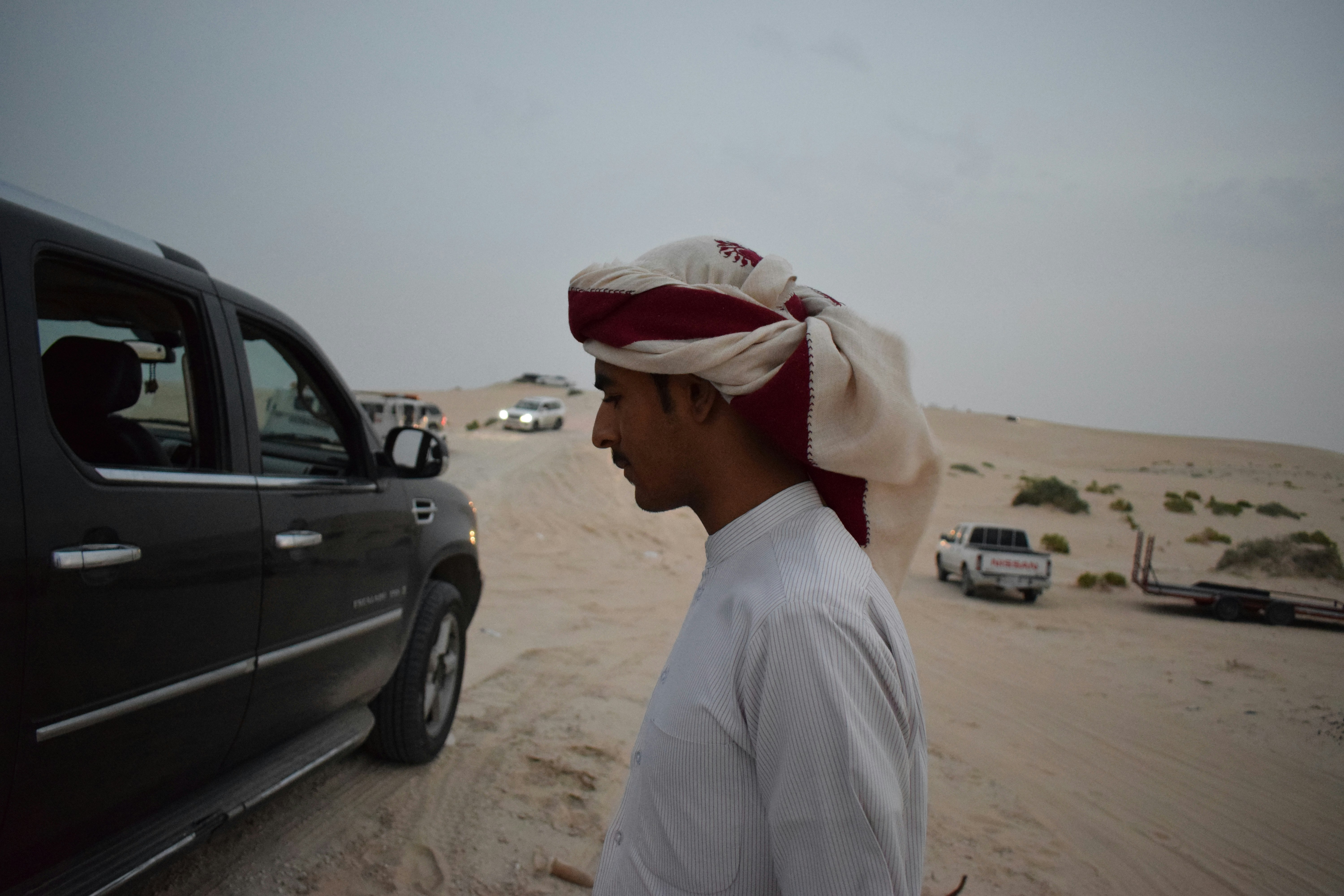 A young man in traditional attire stands beside a vehicle in a desert landscape, capturing the essence of cultural heritage against a backdrop of sand dunes.