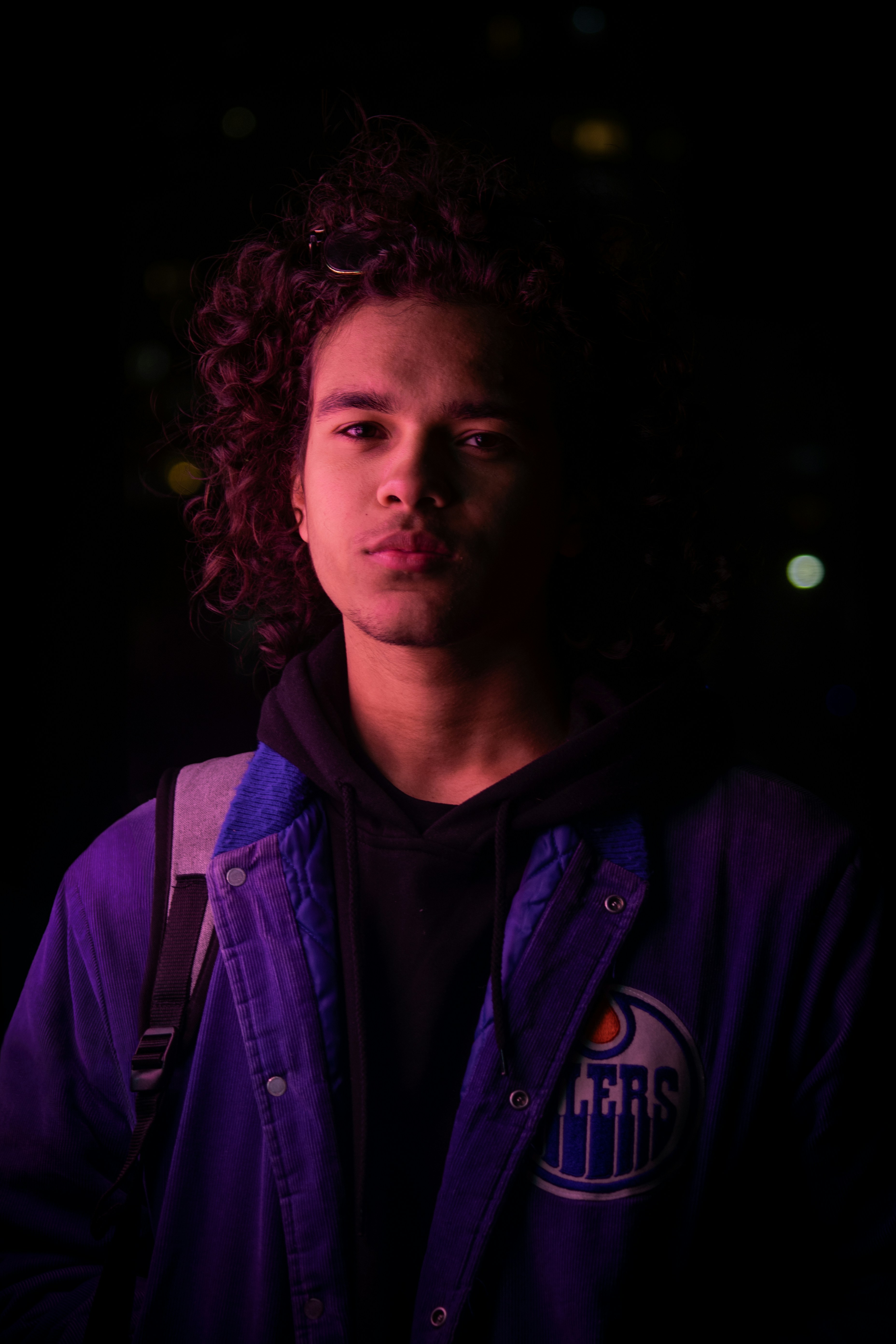 Portrait of a light-skinned man with curly hair standing in front of a pink neon light at night in the winter with a blue jacket and black hoodie