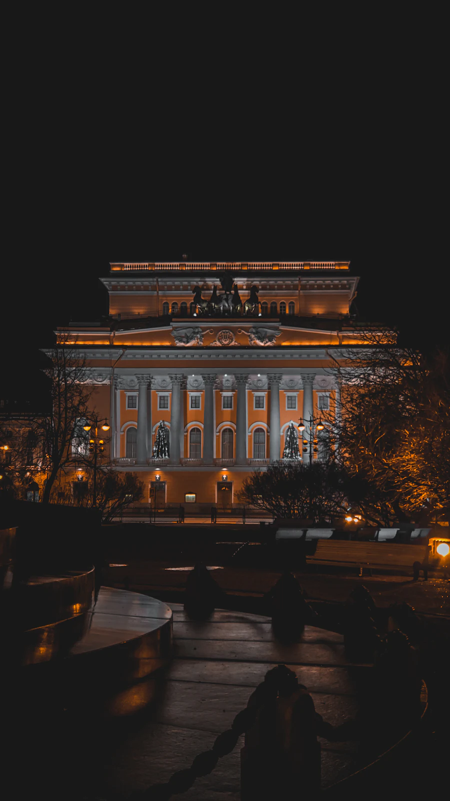 A grand neoclassical building glows with warm orange lights against a dark night sky. Bare trees and dim foreground benches frame the facade.