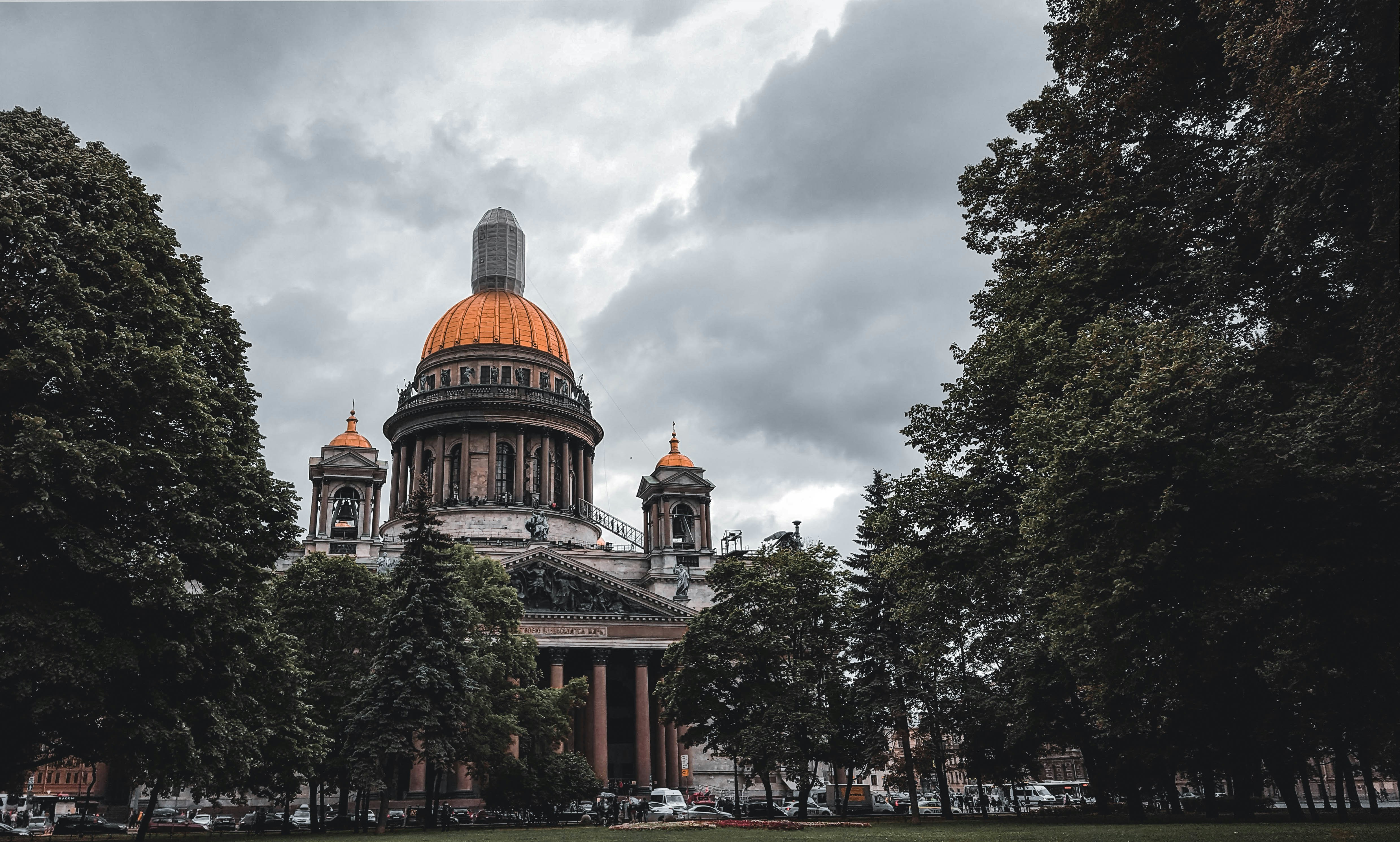 Grand cathedral with a copper dome surrounded by lush greenery against a cloudy sky.