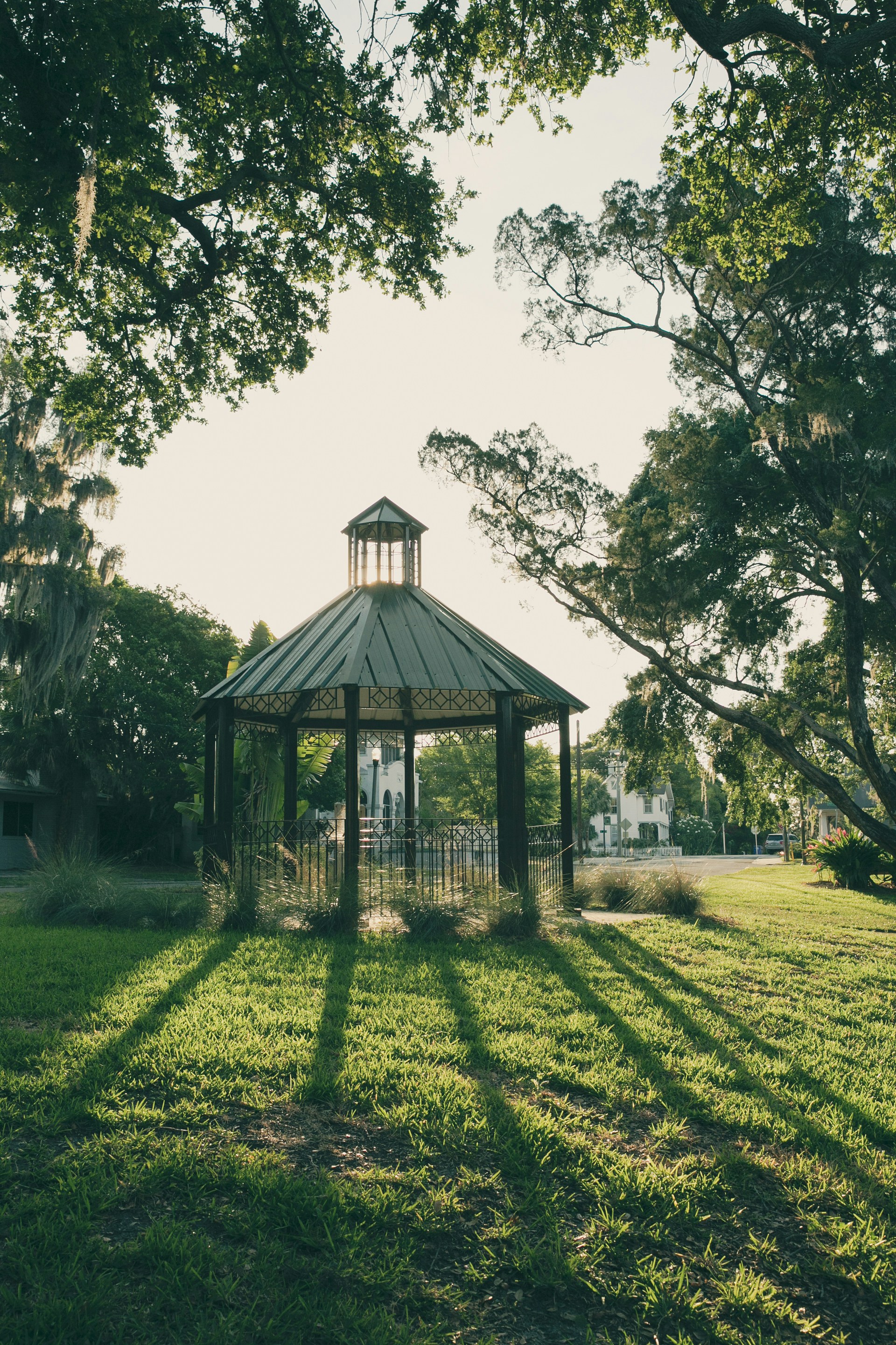 A charming gazebo surrounded by lush greenery, casting long shadows on the grass as the sun sets in the background.
