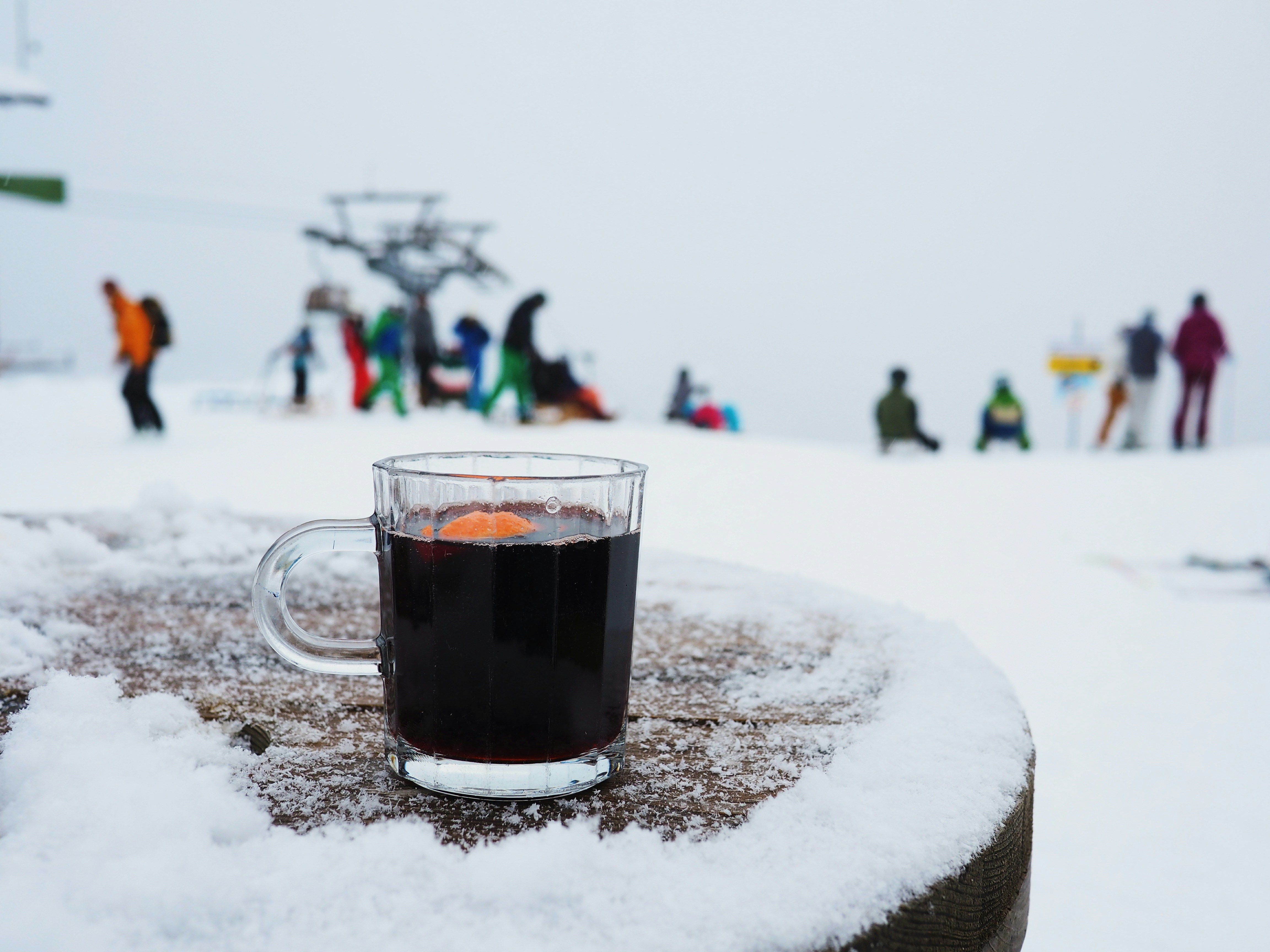 Dark coffee in a glass mug sits on a snow-dusted wooden table, with skiers and bystanders blurred in the wintry background.