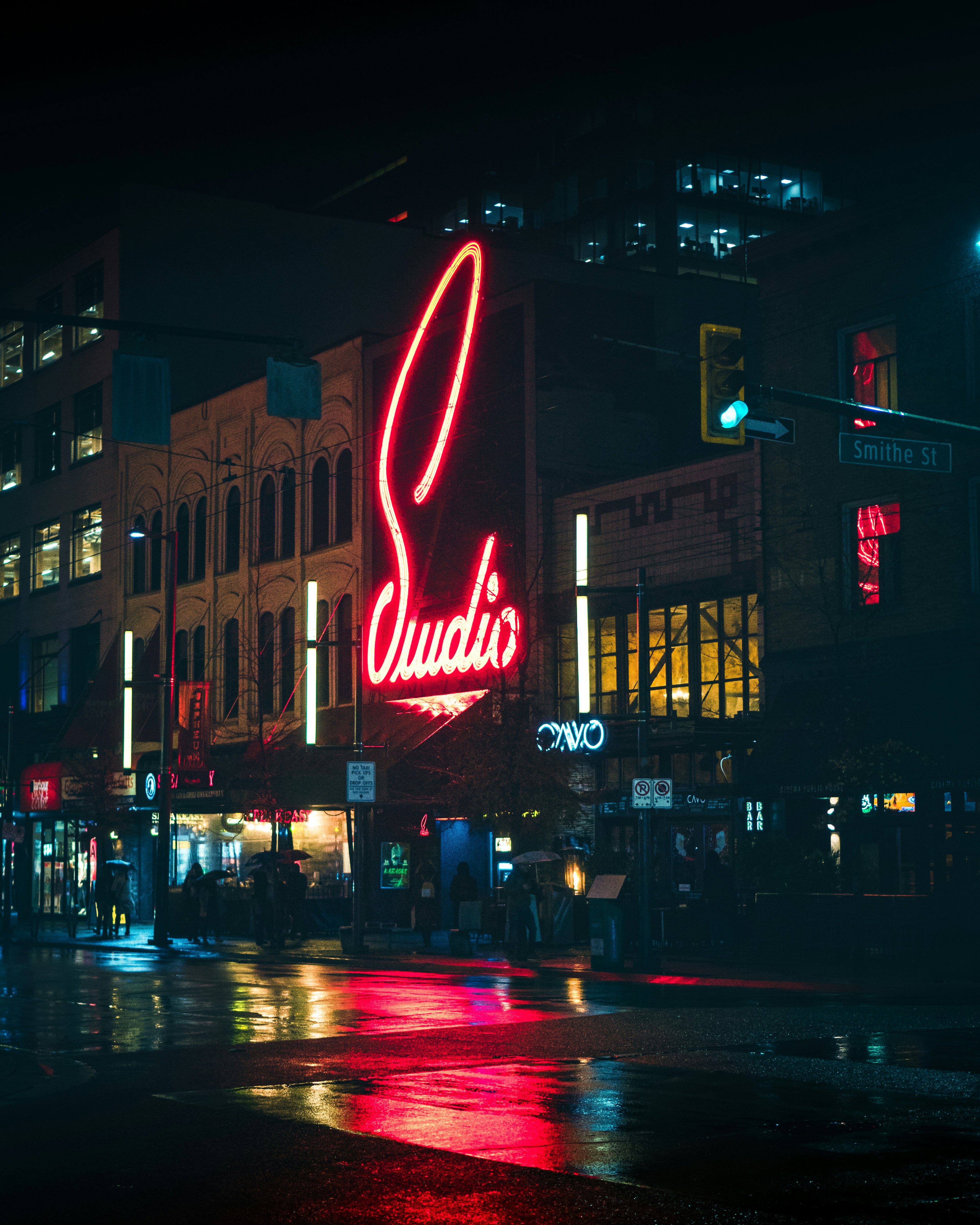 Vibrant neon signage illuminates a bustling street corner at night, reflecting off the wet pavement. The scene captures the lively atmosphere of urban nightlife.