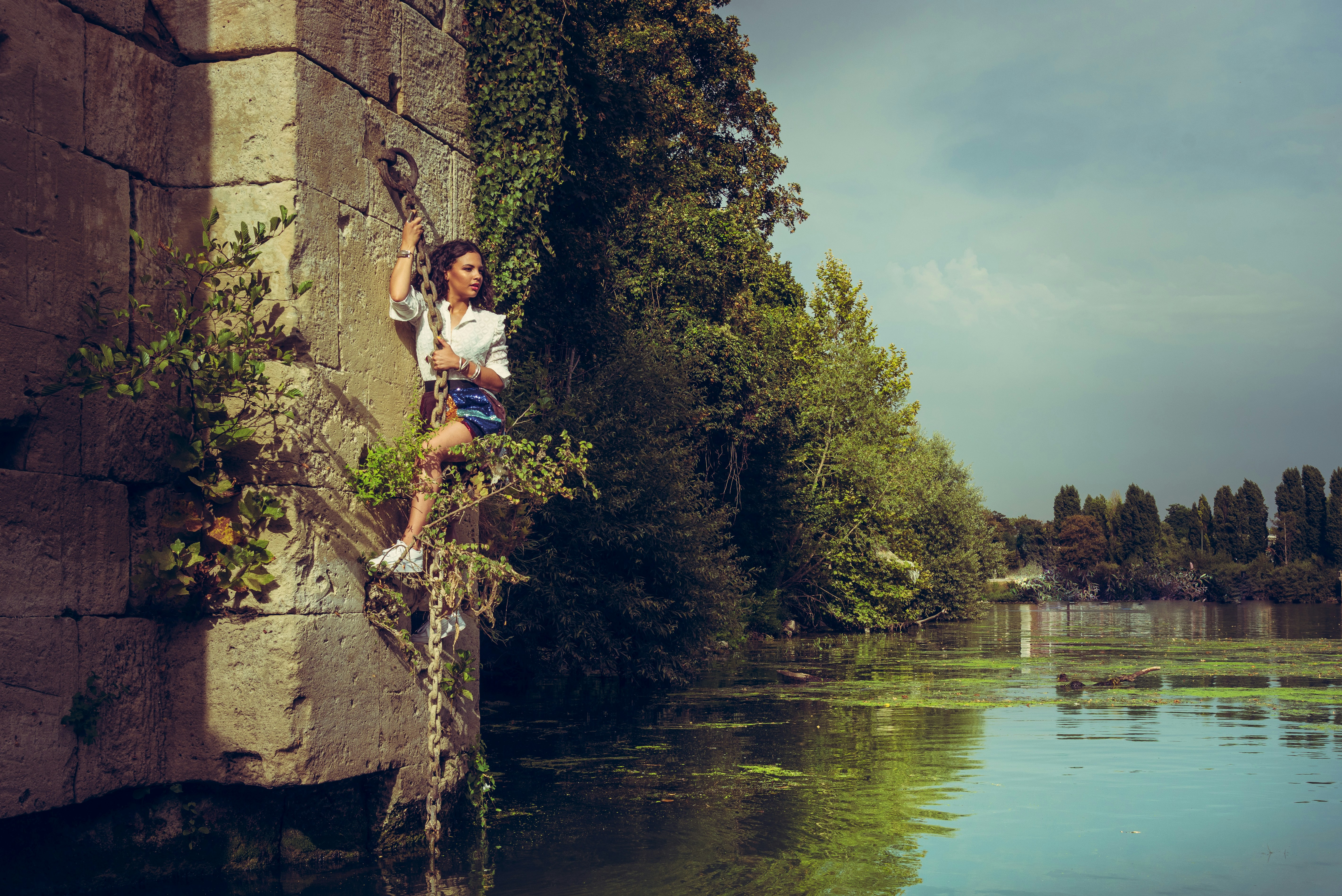 Riverside Ascent: Climber Clings to a Mossy Wall Over Calm Waters