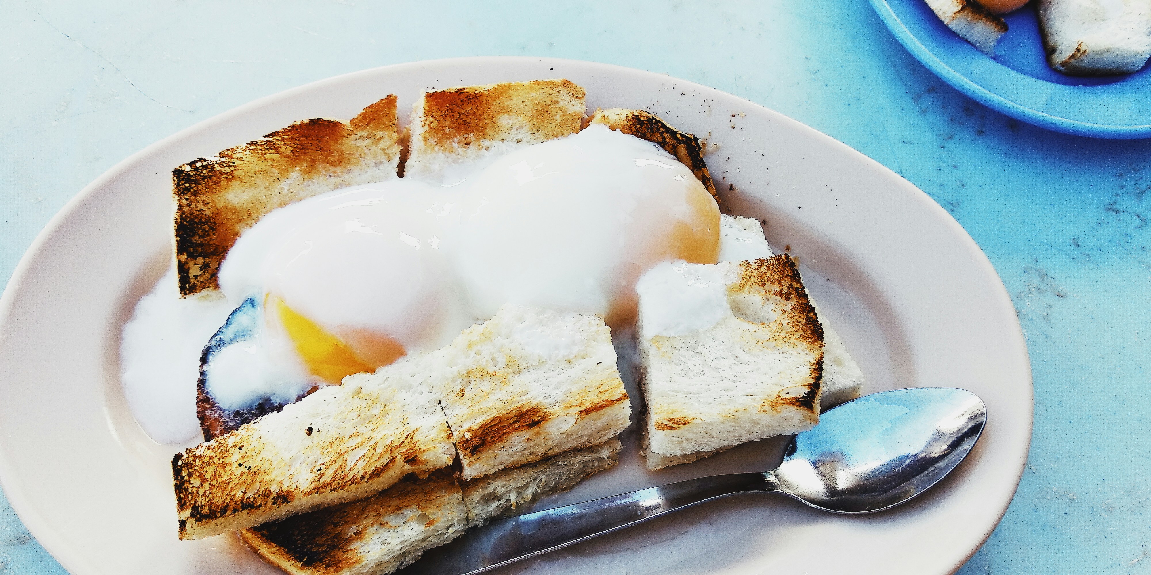 Soft-boiled eggs resting on toasted bread, served on a pale plate with a silver spoon beside it. A blue plate with additional toast is visible in the background.