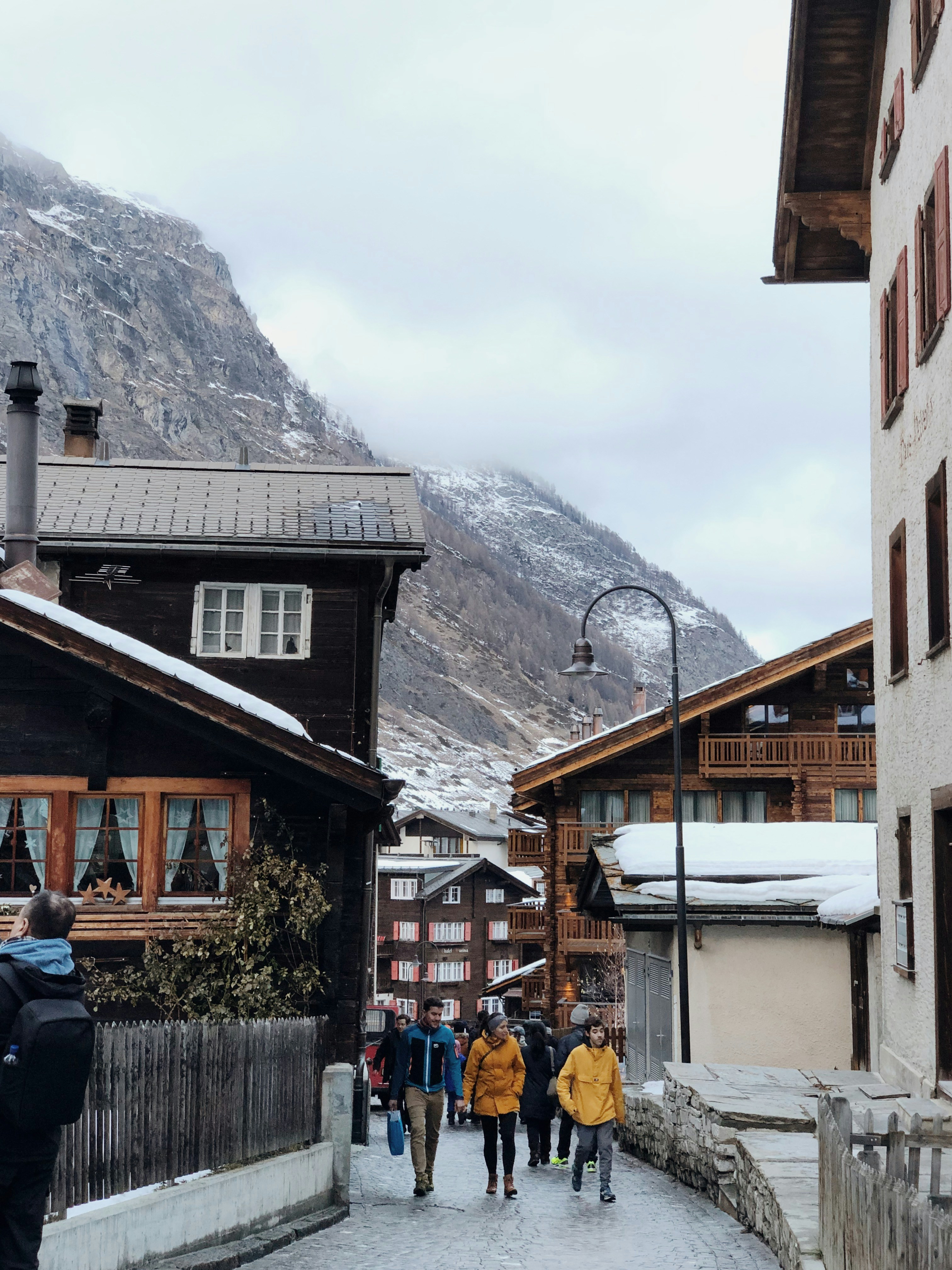 Visitors meander through a quaint, snow-covered village lined with traditional wooden chalets and majestic mountains in the background.