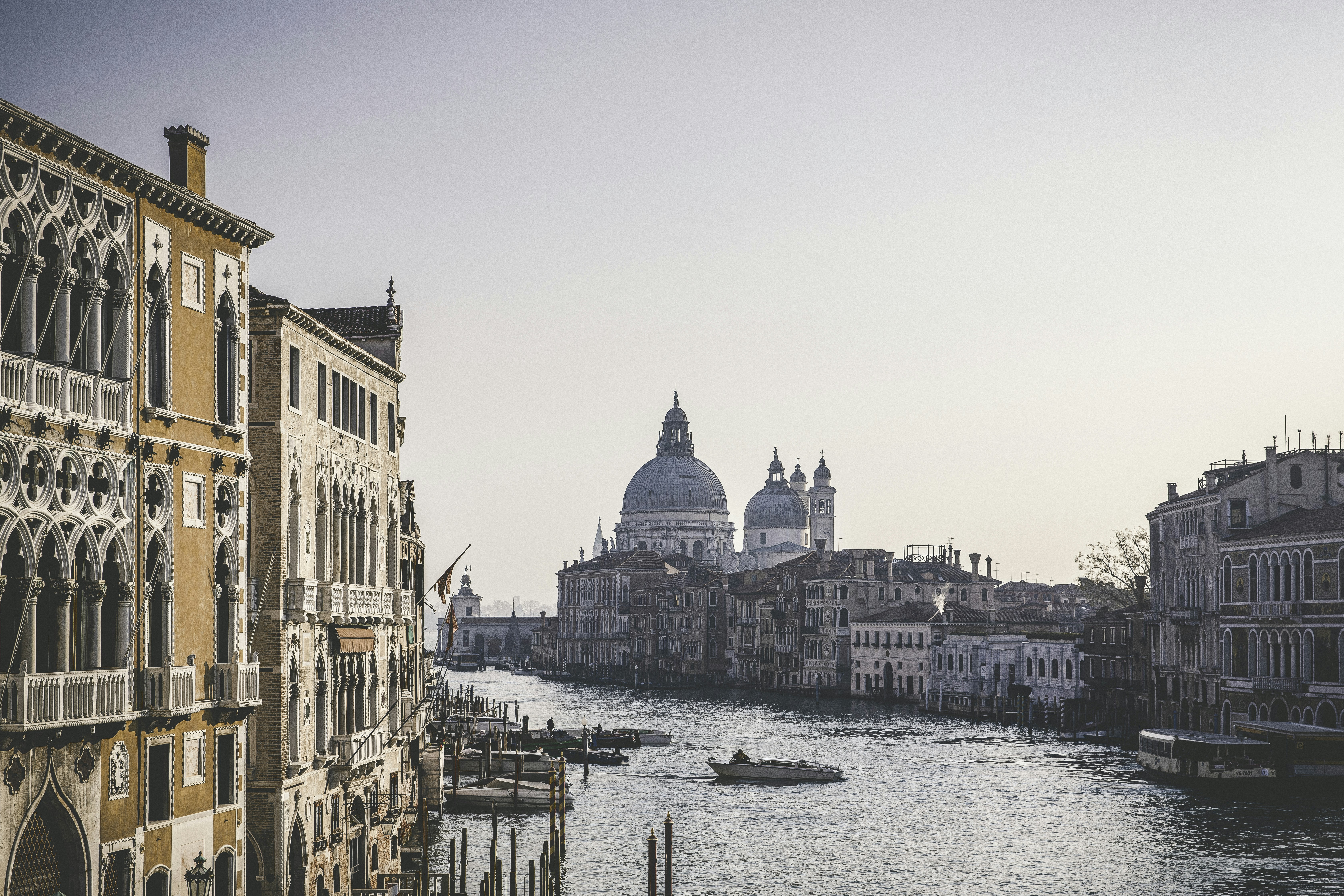 This captivating image showcases the tranquil beauty of Venice's Grand Canal under the soft morning light. The composition features the iconic architecture of the city, with intricate facades in warm earth tones lining the canal, leading the eye toward the majestic dome in the distance. The gentle lighting creates a peaceful atmosphere, highlighting the subtle blues and greys of the water, making the image both visually striking and evocative of Venice's timeless charm.