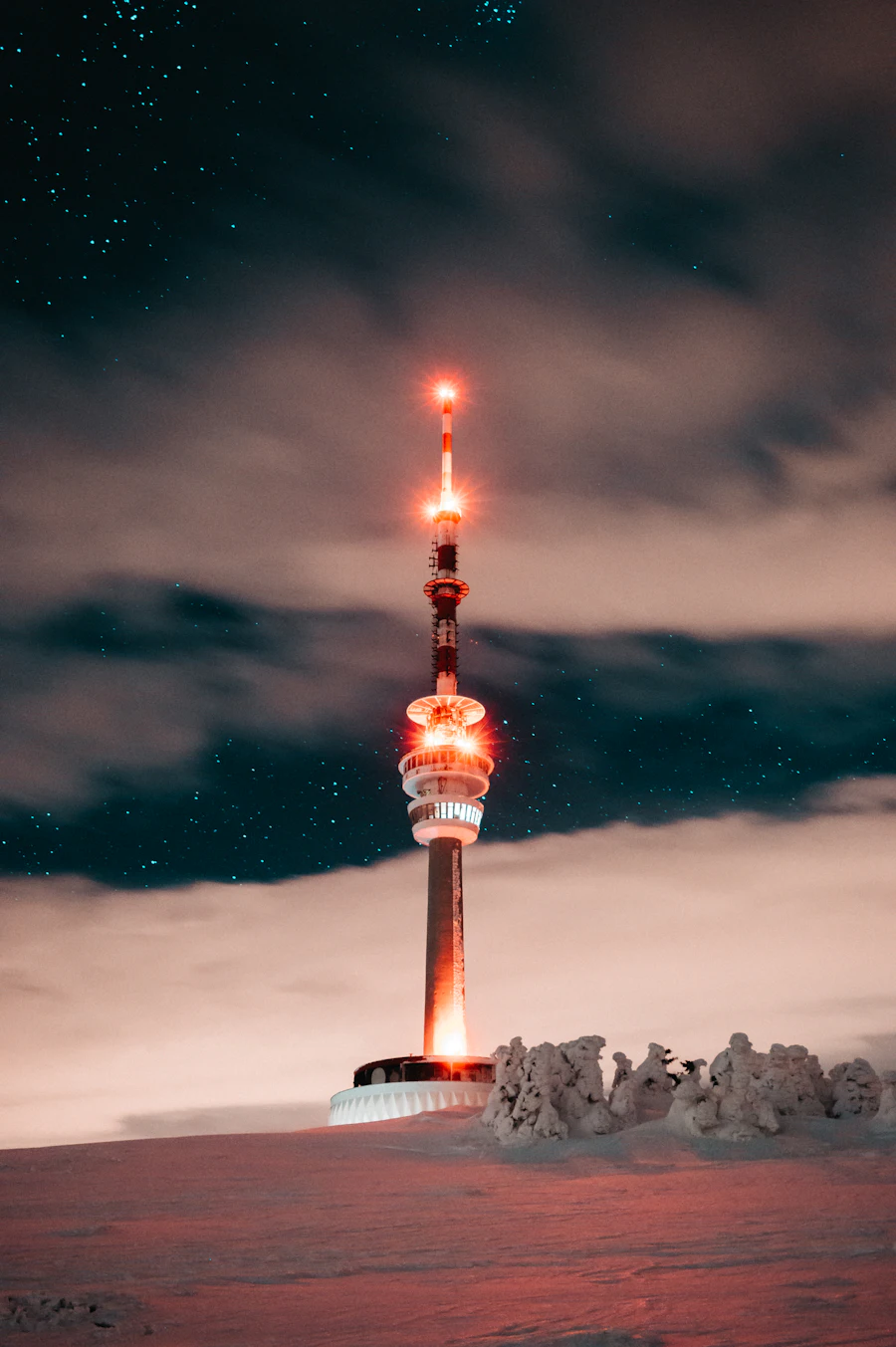 A tall communications tower glows red against a cloudy night sky dotted with stars, rising from a snowy landscape. Orange lights reflect off the snow and nearby frost-covered rocks.