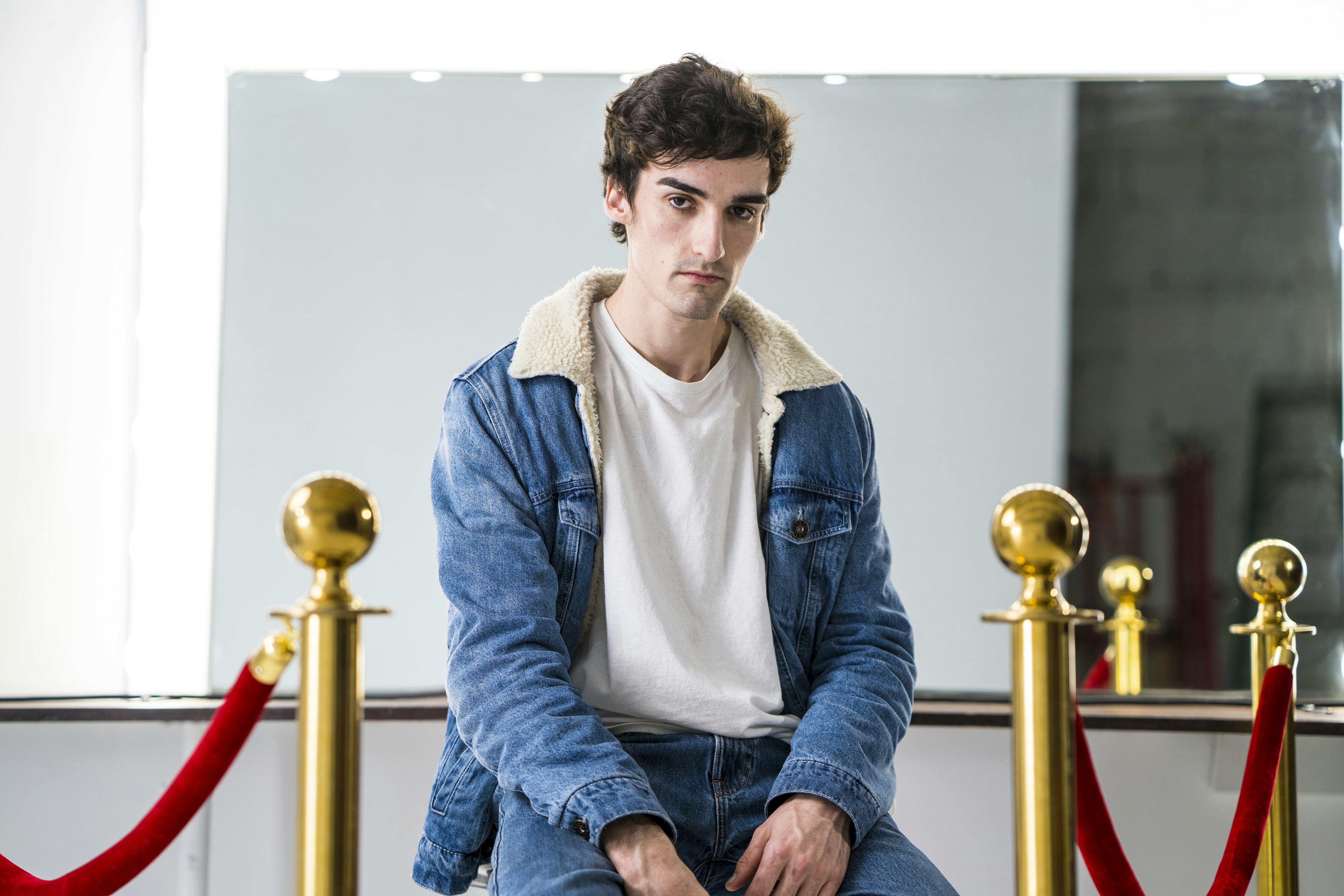 Man wearing blue and white denim jacket sitting near brass stanchion ...