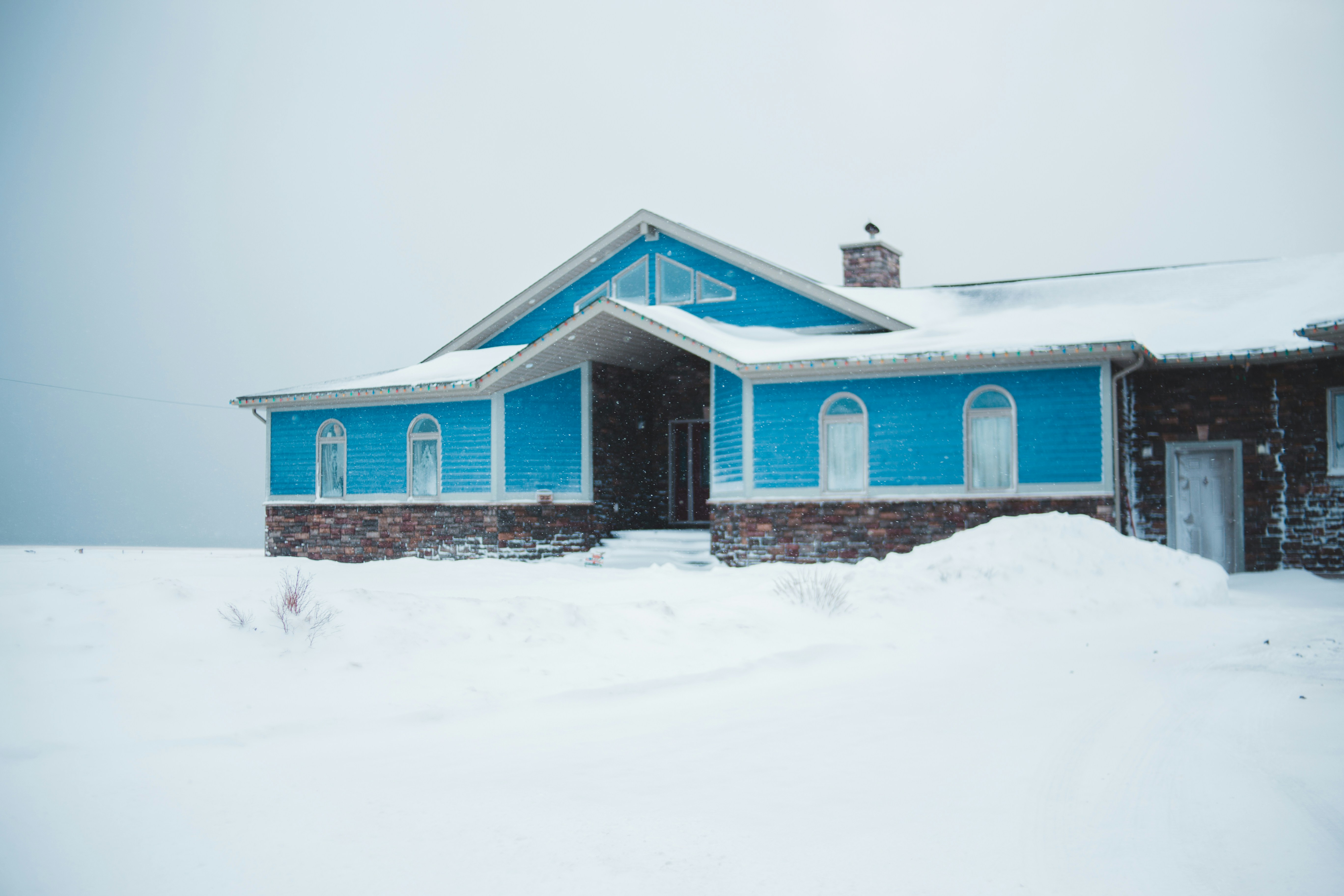 A charming blue house stands resilient against a backdrop of falling snow, surrounded by a pristine white landscape.