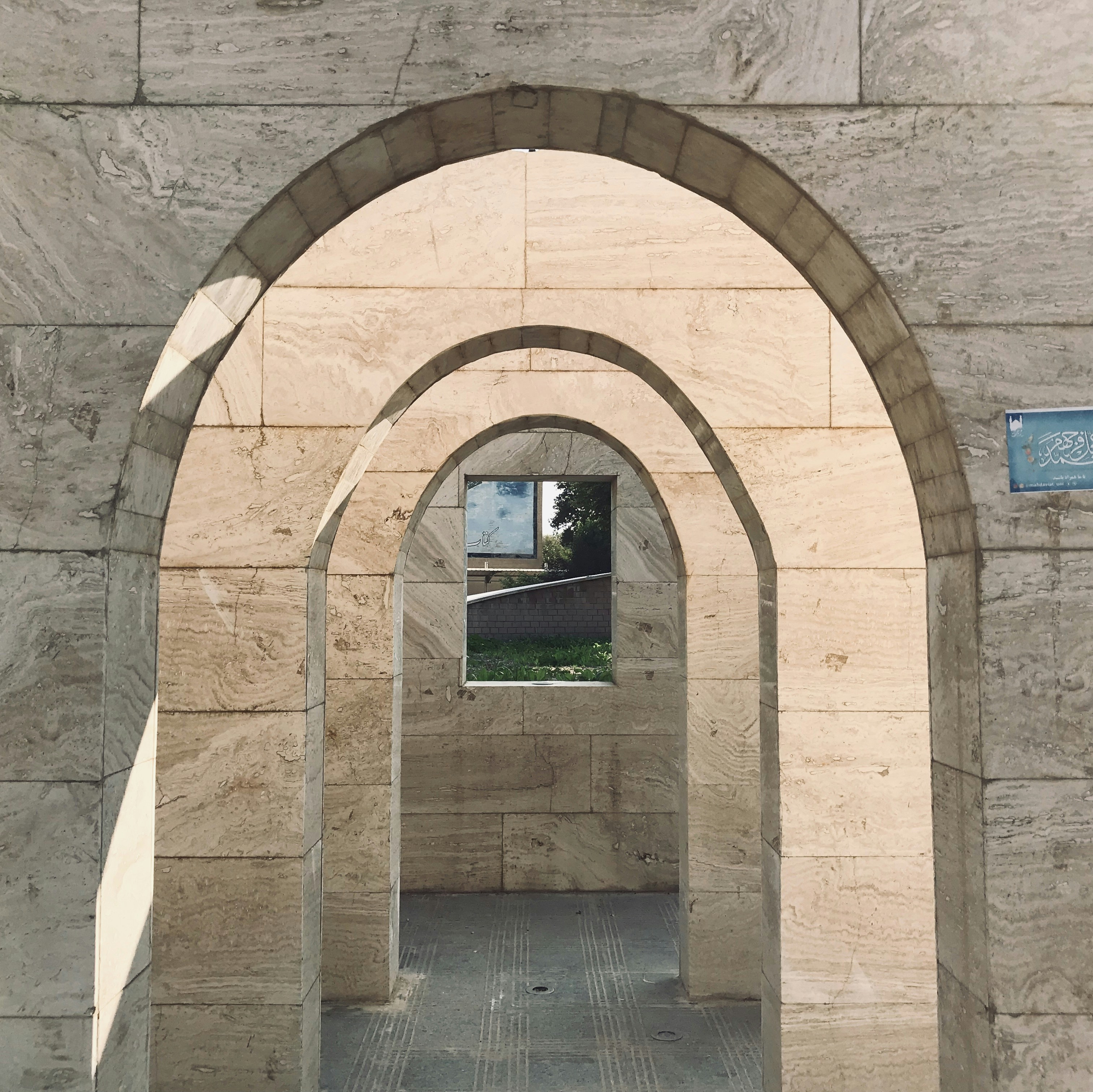 Three elegantly arched doorways framed in textured stone, leading to a glimpse of greenery beyond. A sign hangs to the right, adding context to the scene.