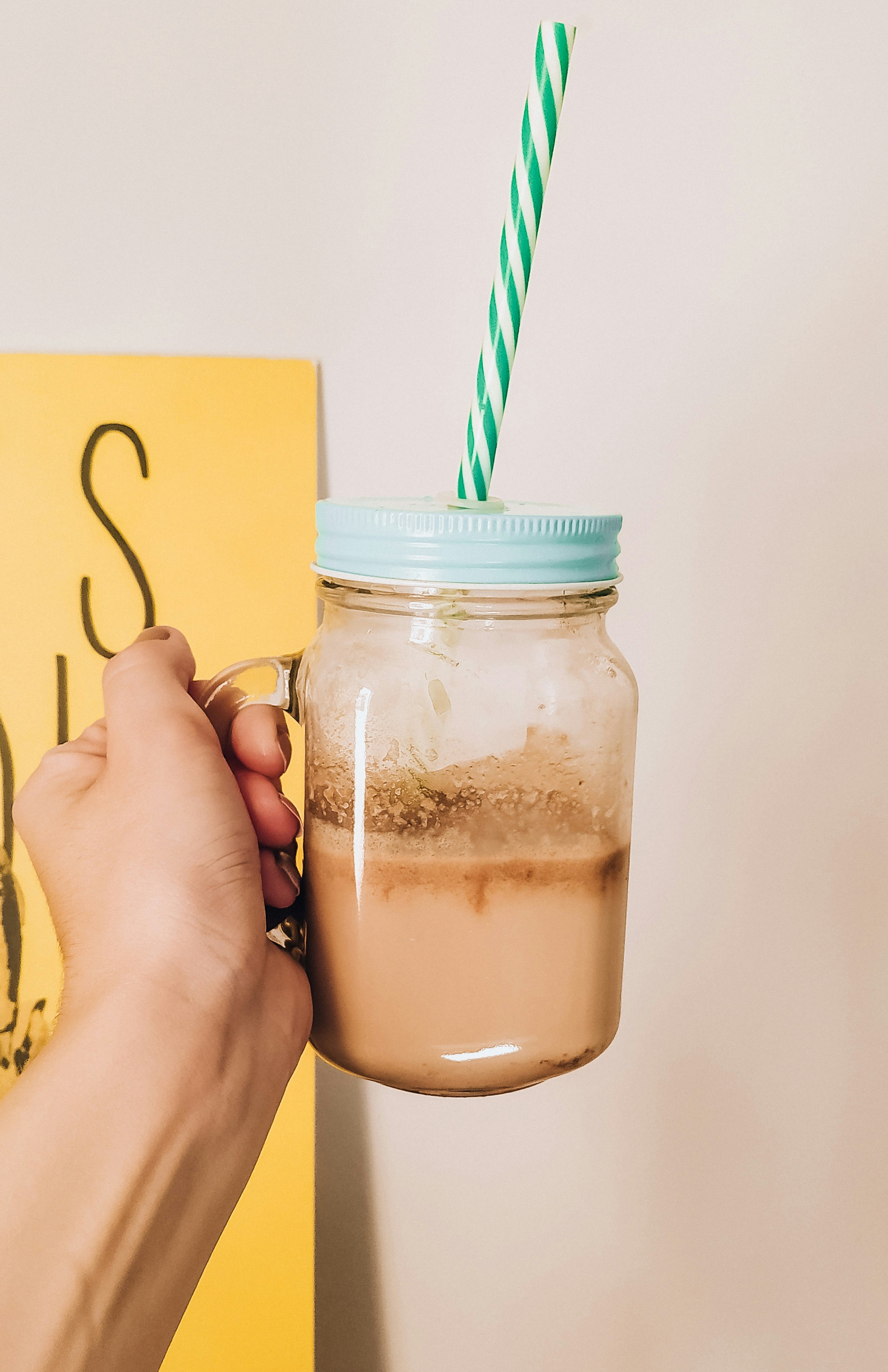 A hand holds a mason jar filled with a creamy beverage, featuring a striped straw, against a vibrant yellow backdrop with artistic lettering.