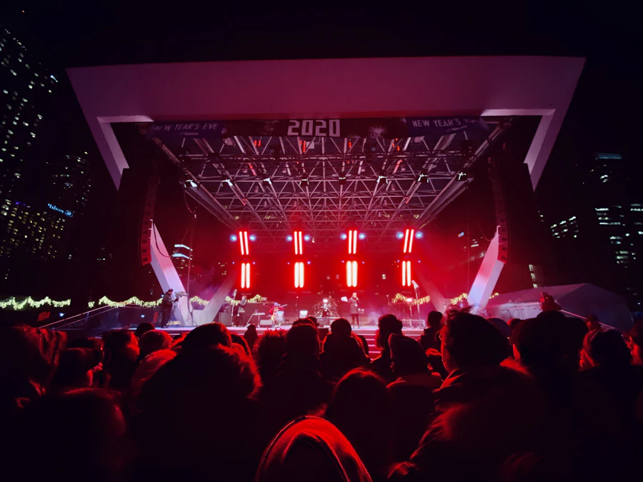 Crowd gathered in front of a large outdoor concert stage glowing red under a modern canopy. Bright vertical light bars frame the performers with city buildings visible in the background.