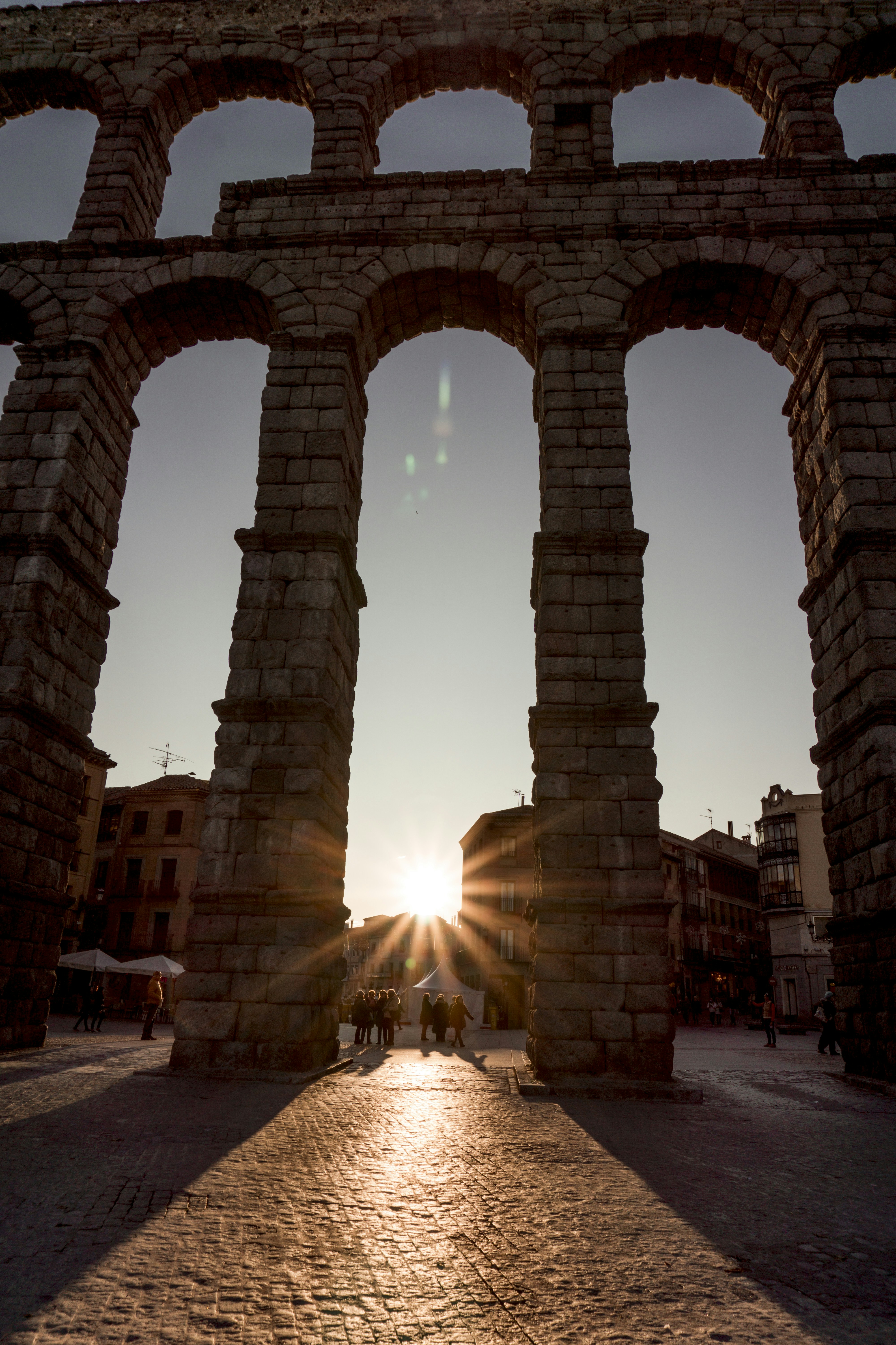 Sun setting behind a stone aqueduct, casting long shadows on the cobblestones.