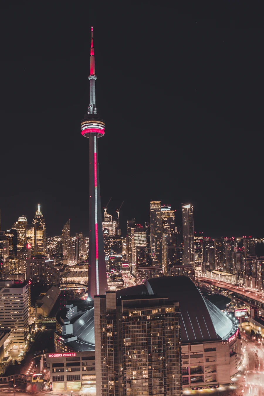 The CN Tower rises above the illuminated Toronto skyline, with Rogers Centre in the foreground and city lights stretching into the distance at night.