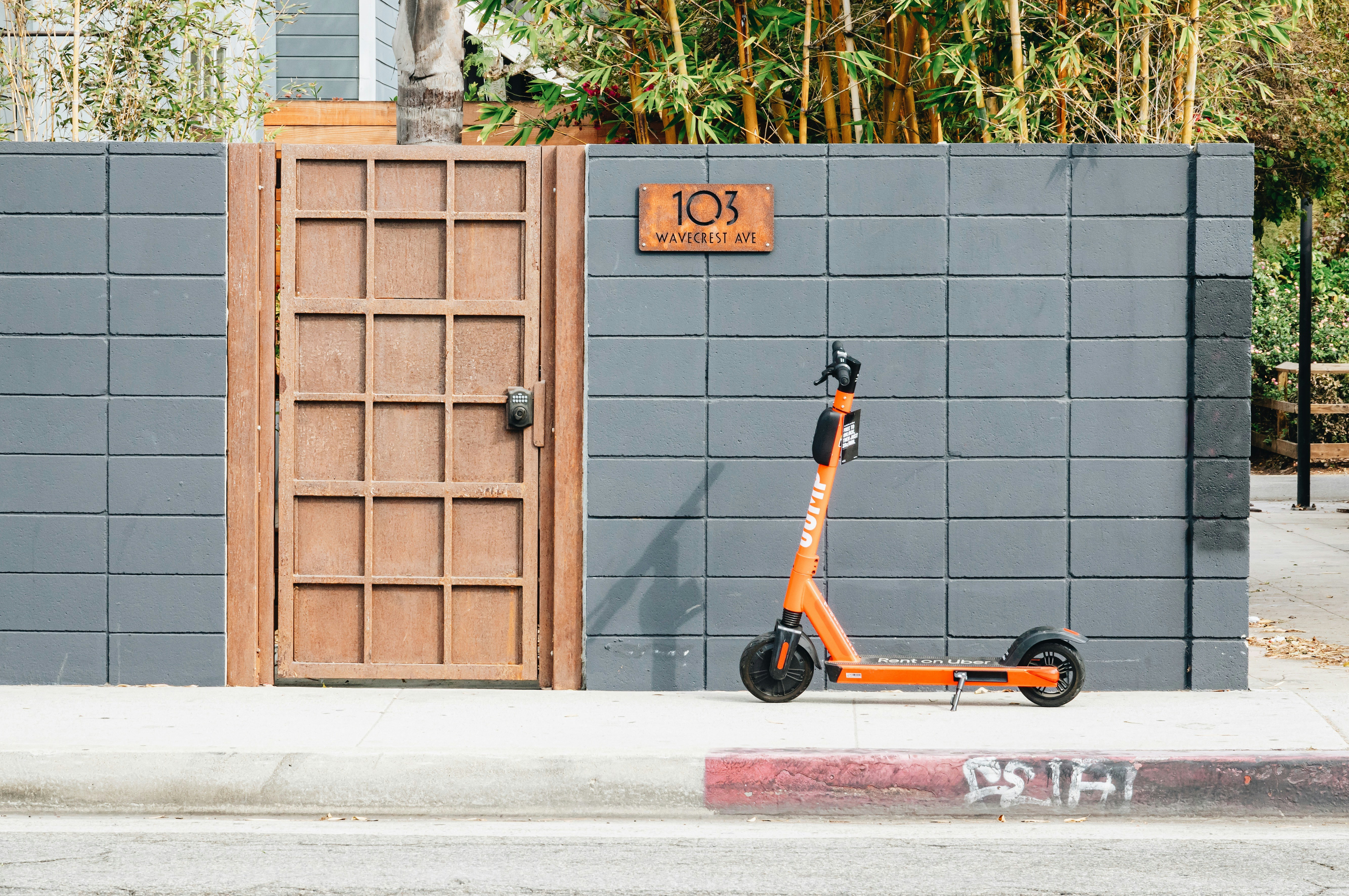An orange electric scooter parked beside a textured gray wall featuring a wooden door and a house number plaque. The scene captures a blend of modern transportation and architectural detail.