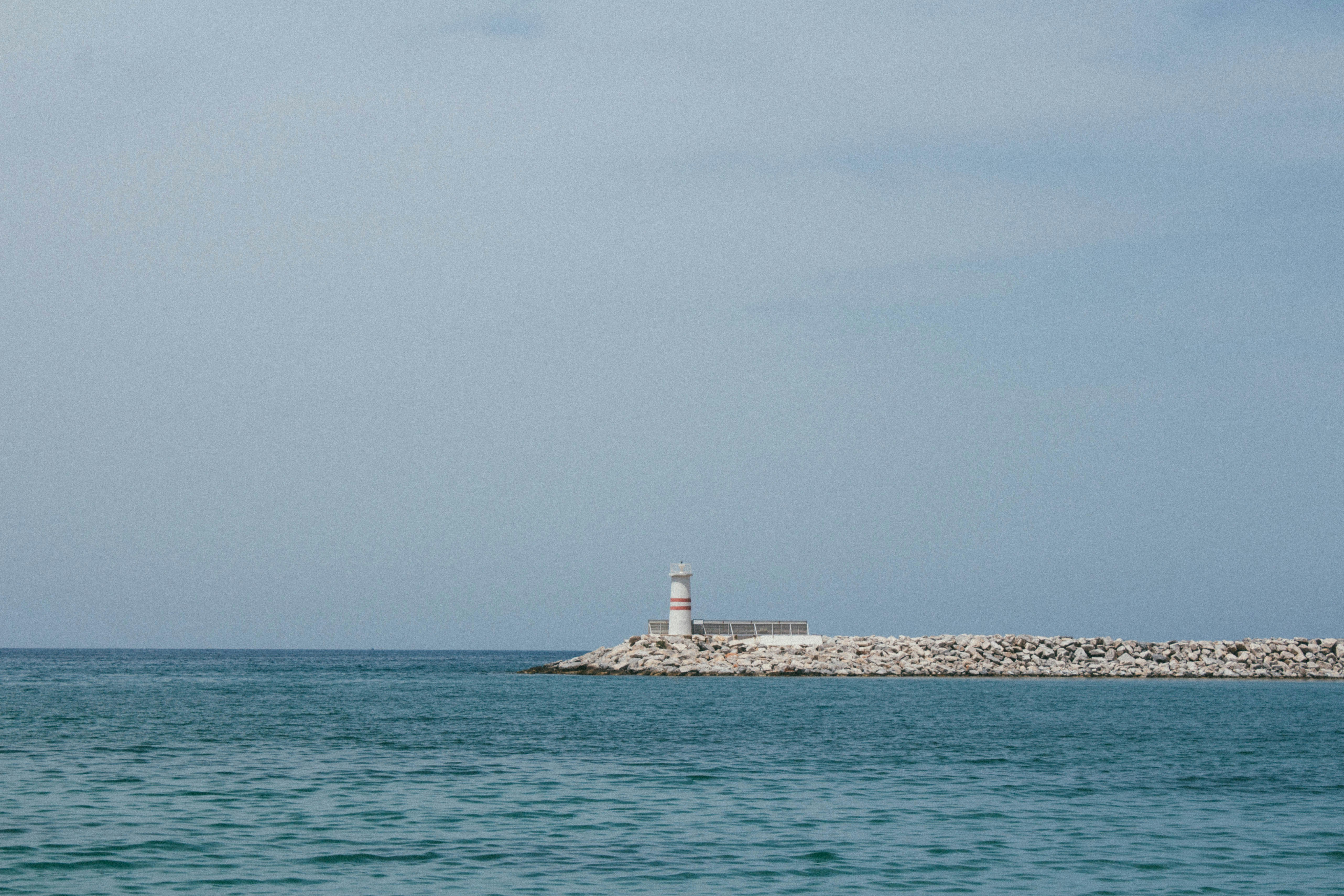 A lighthouse stands resilient on a rocky pier, overlooking a serene ocean under a clear sky.