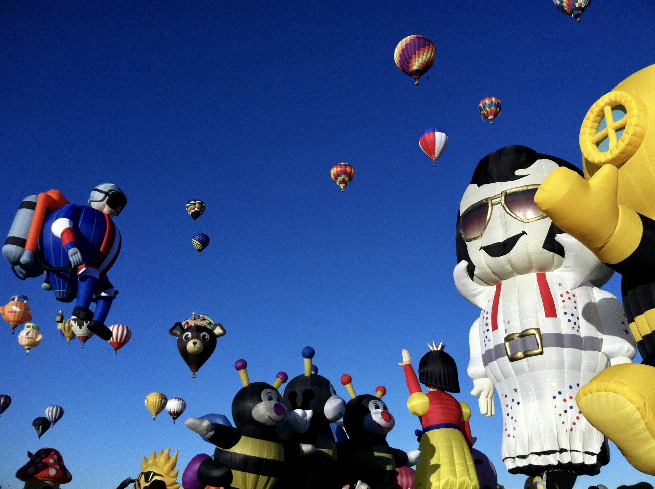 Colorful hot air balloons in unique shapes and designs float against a clear blue sky.