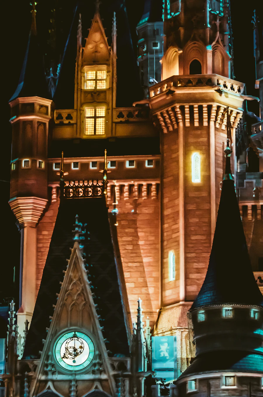 Nighttime view of a glowing fairytale castle with turrets, arched windows, and a bright clock face in the foreground. Warm amber lighting contrasts with teal accents against the dark sky.