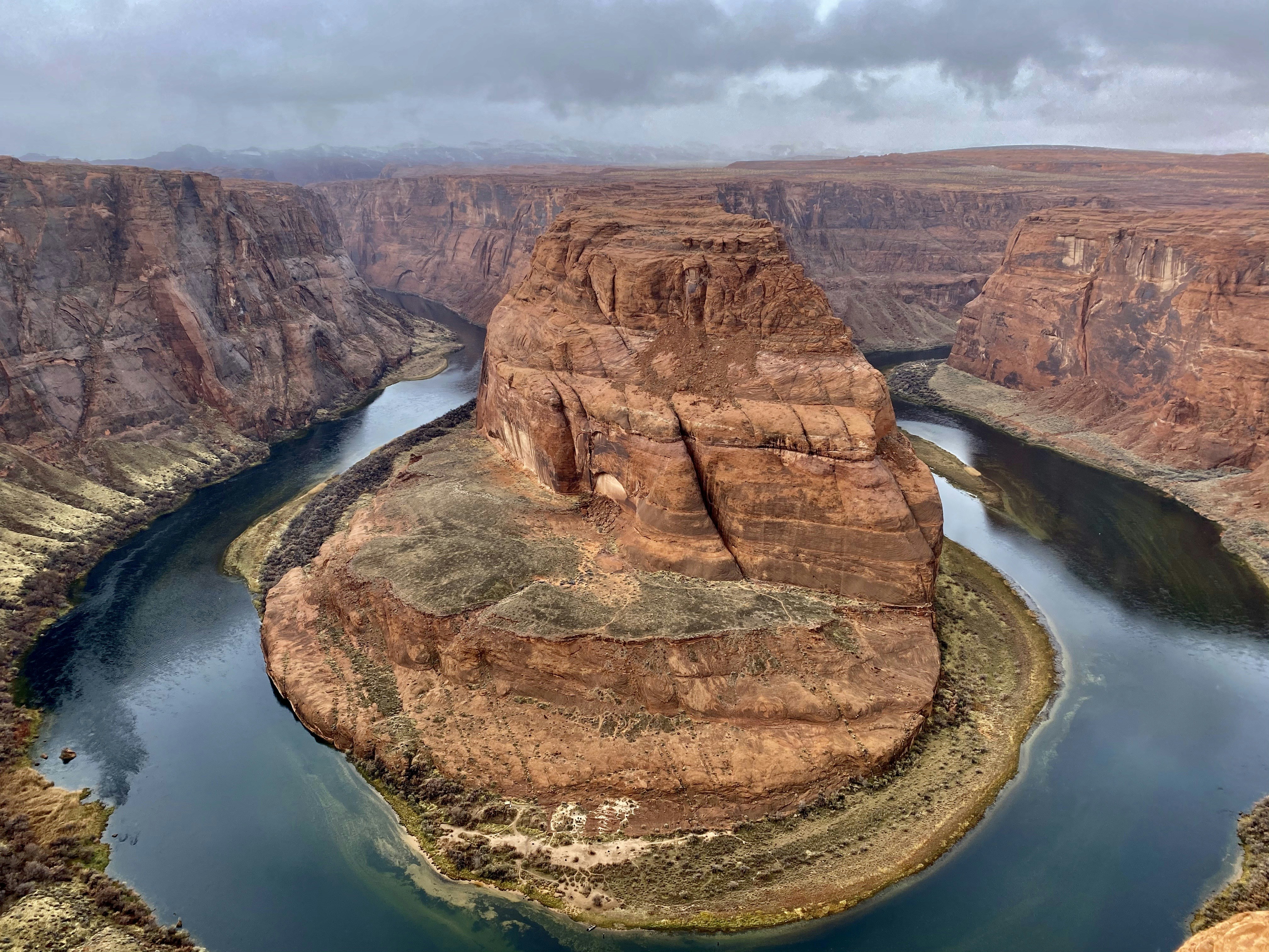 Aerial view of Horseshoe Bend, showcasing the dramatic curves of the Colorado River winding around a sandstone formation. The rugged terrain contrasts with the reflective water below.