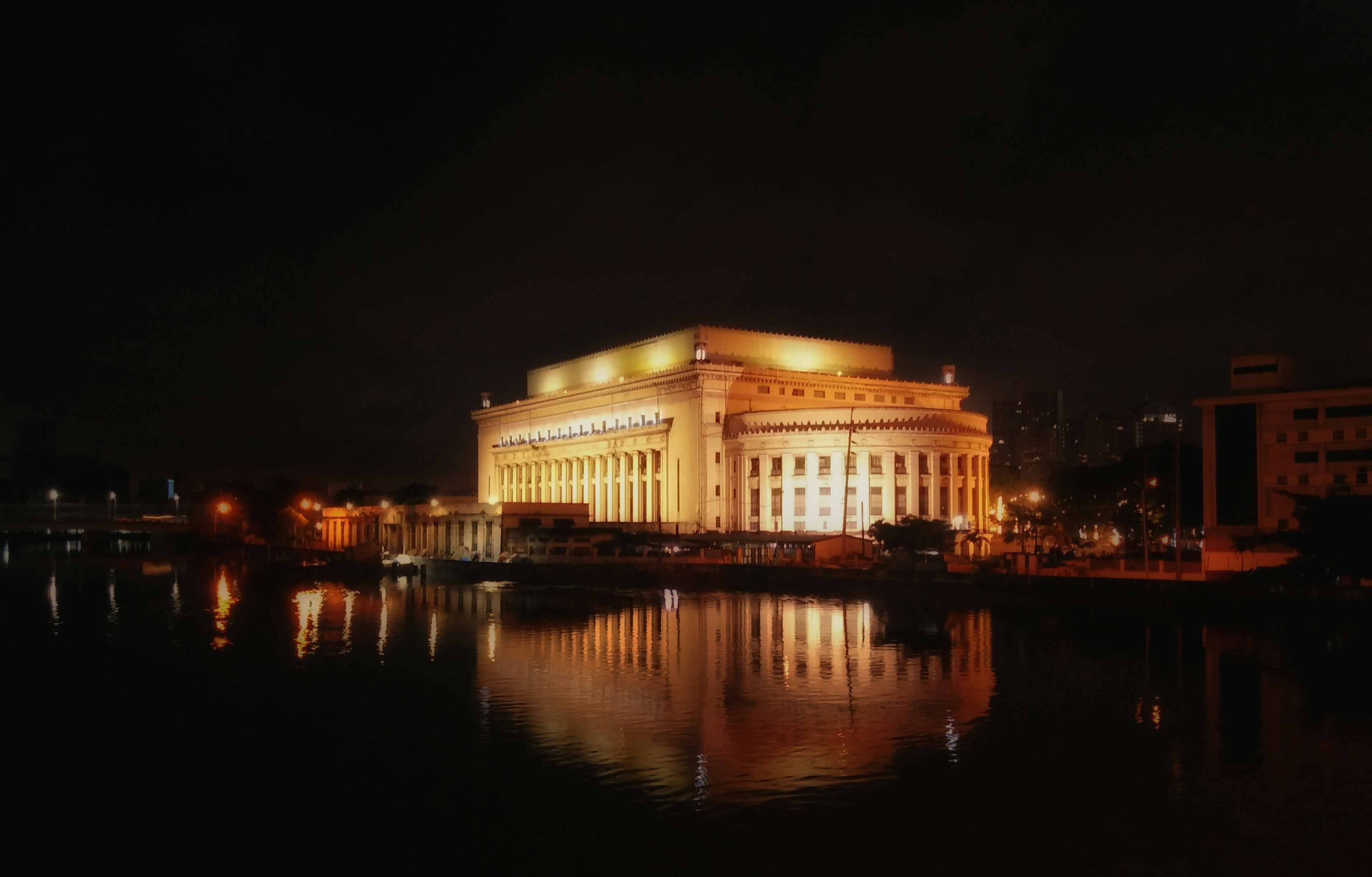 Night photograph of a grand neoclassical hall lit in warm tones, reflected in the calm water.
