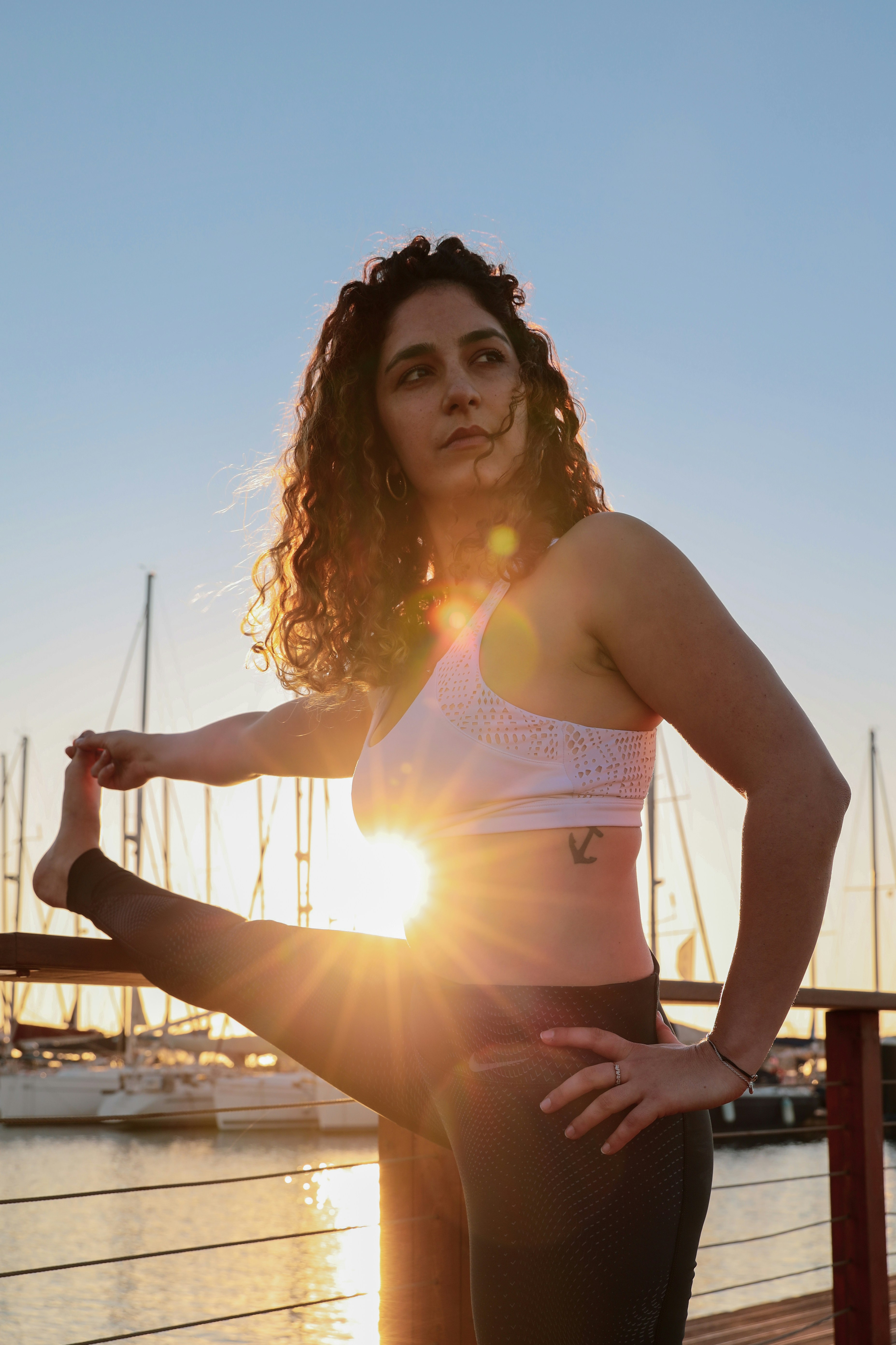 Woman performing a yoga stretch on a dock at sunset, with sailboats in the background and sunlight creating a halo effect.