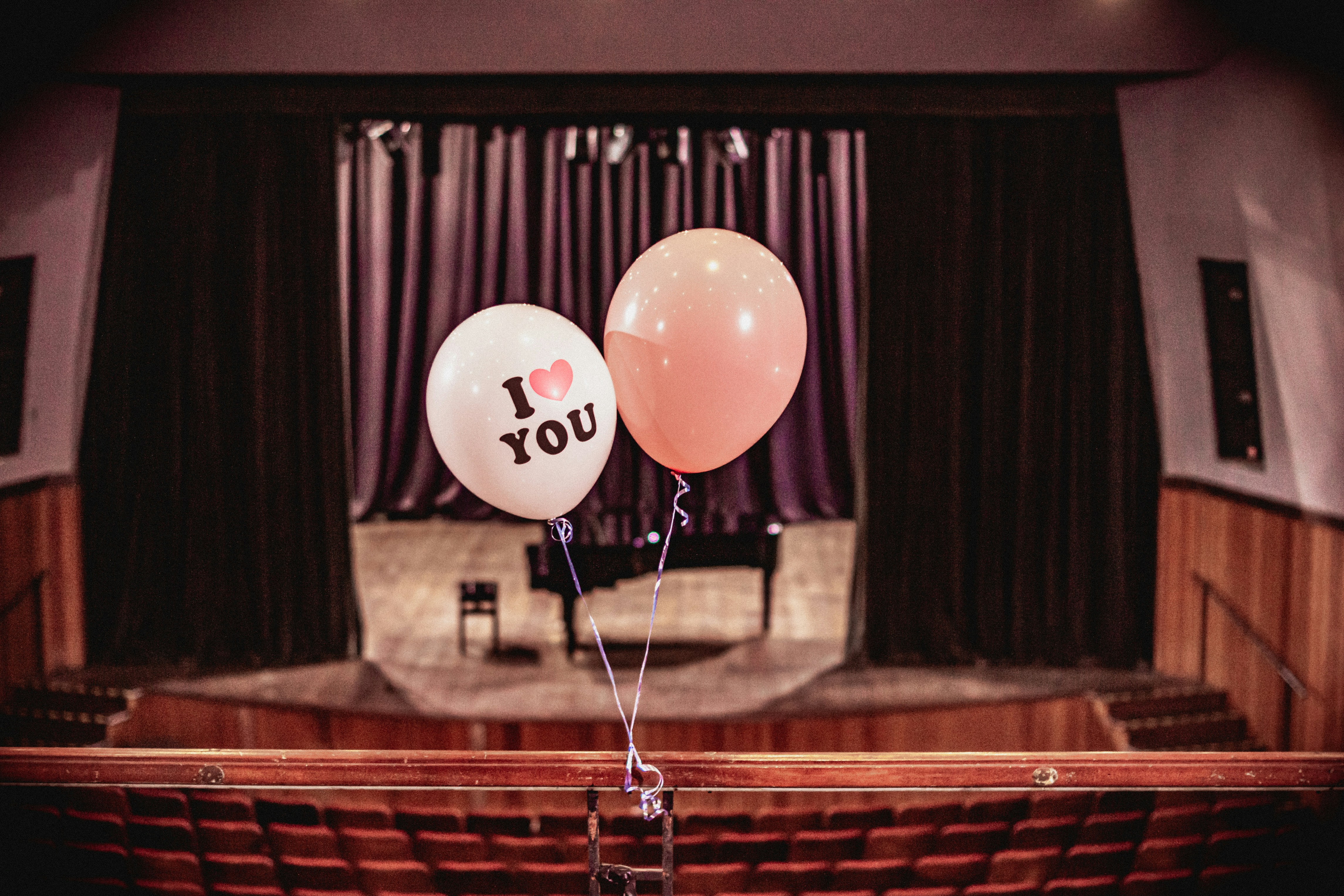 Two balloons, one with 'I ♥ YOU,' float in front of a stage with a piano and dark curtains.