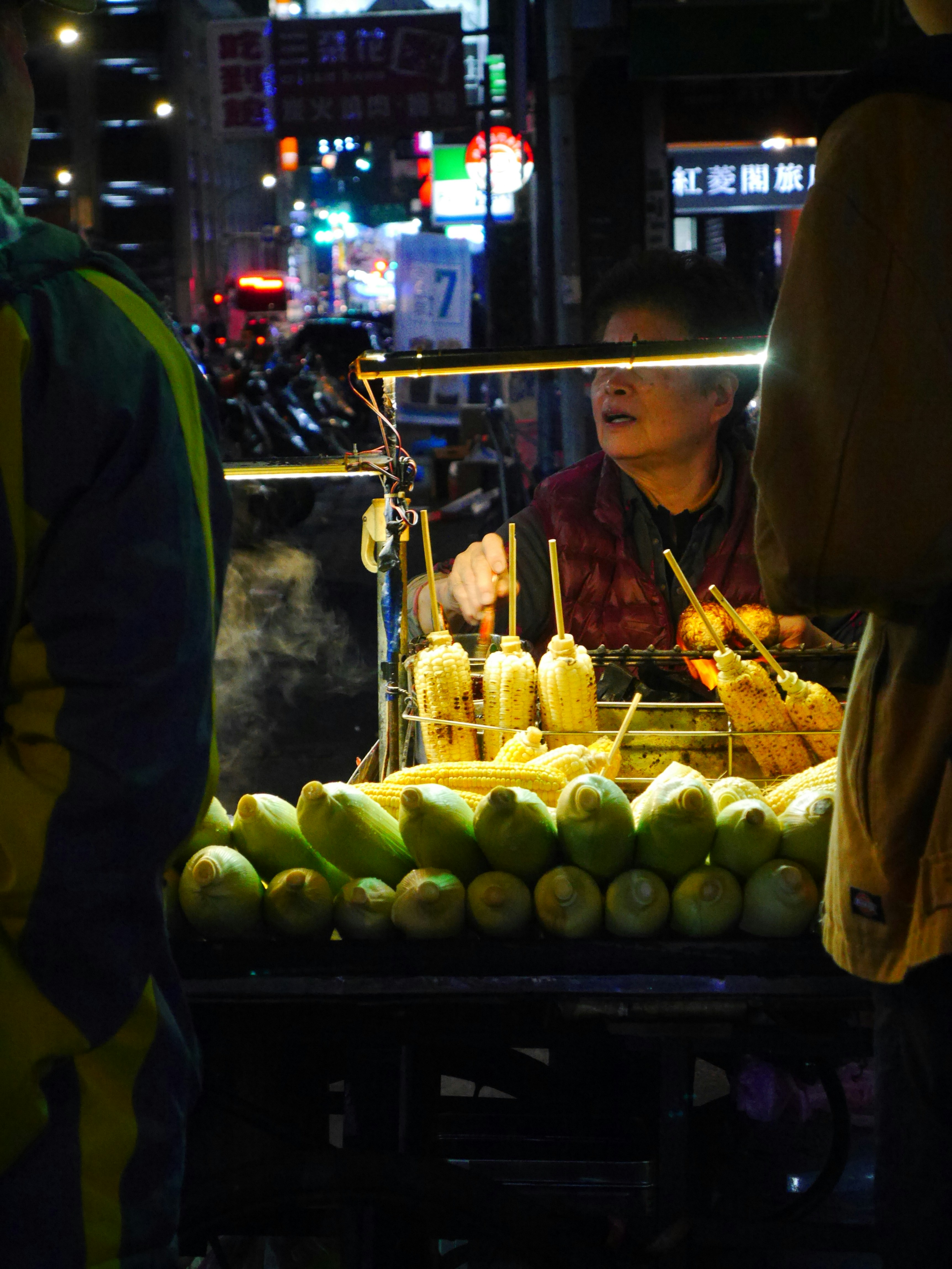 Night market stall with grilled corn and skewered treats under neon lights, a vendor's face illuminated by a horizontal lamp.