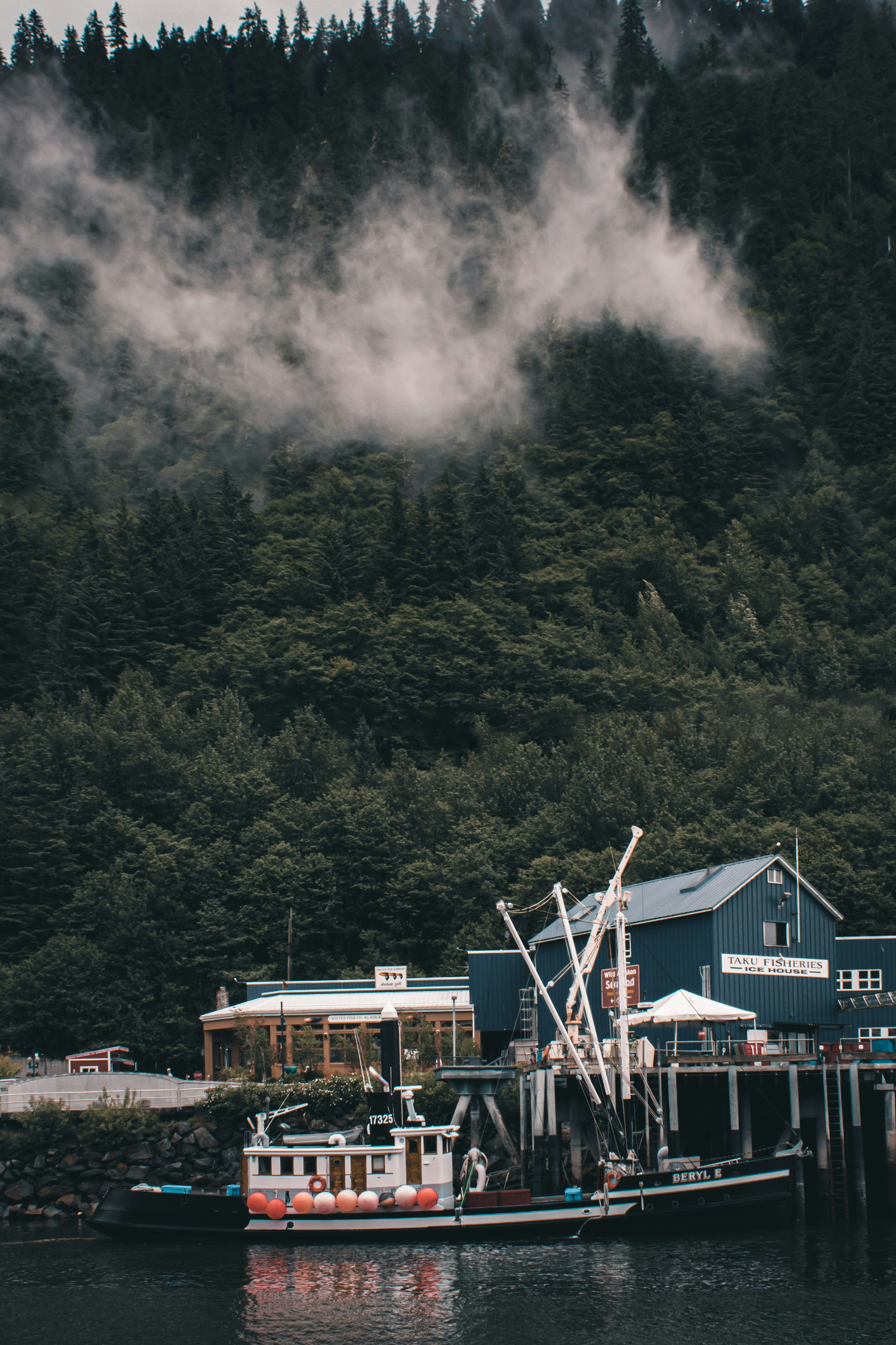 Fishing boat docked beside a blue and white building with a forested hillside shrouded in mist.
