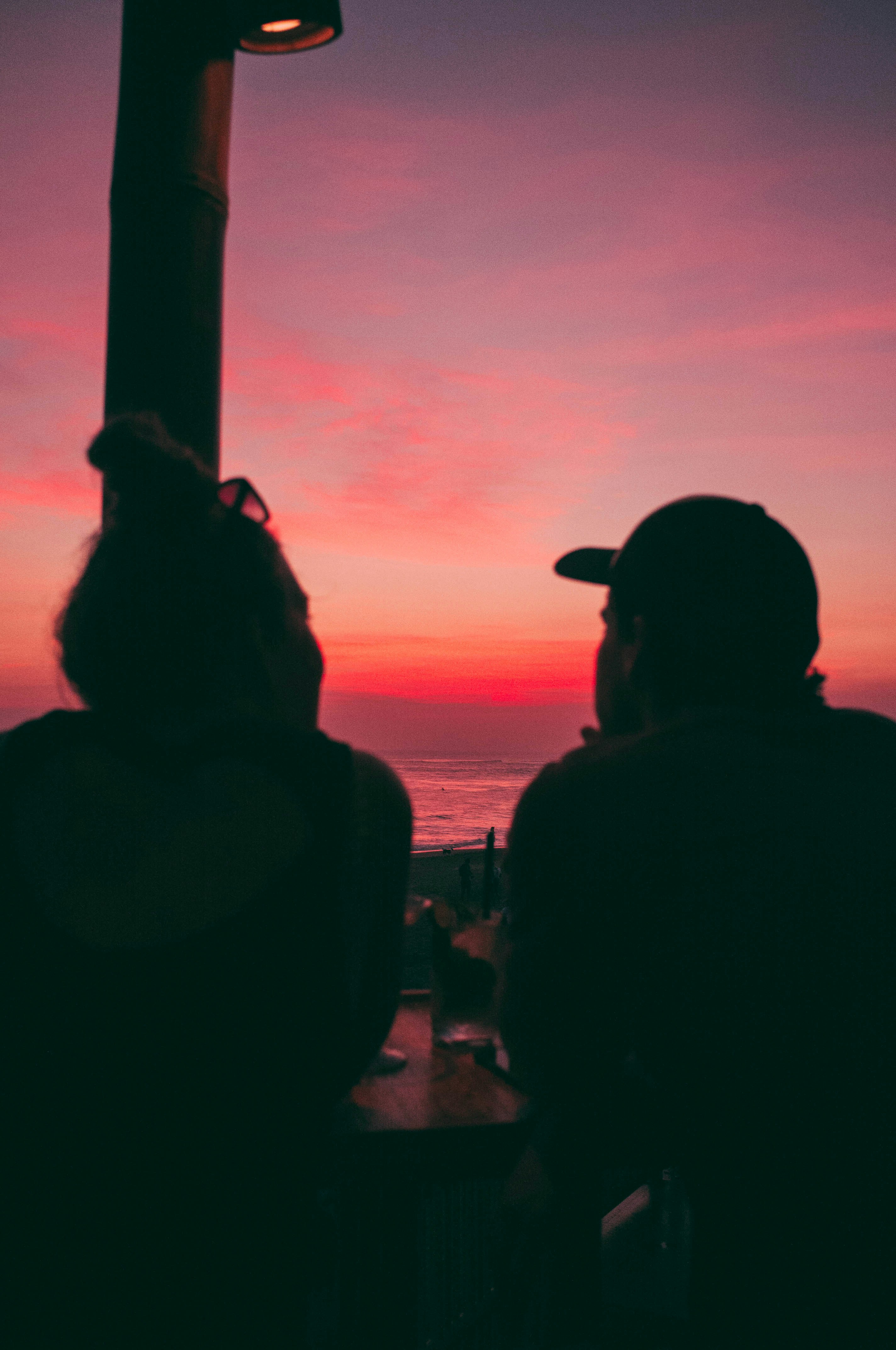 Two people silhouetted against a vibrant pink and orange sunset, overlooking the ocean.